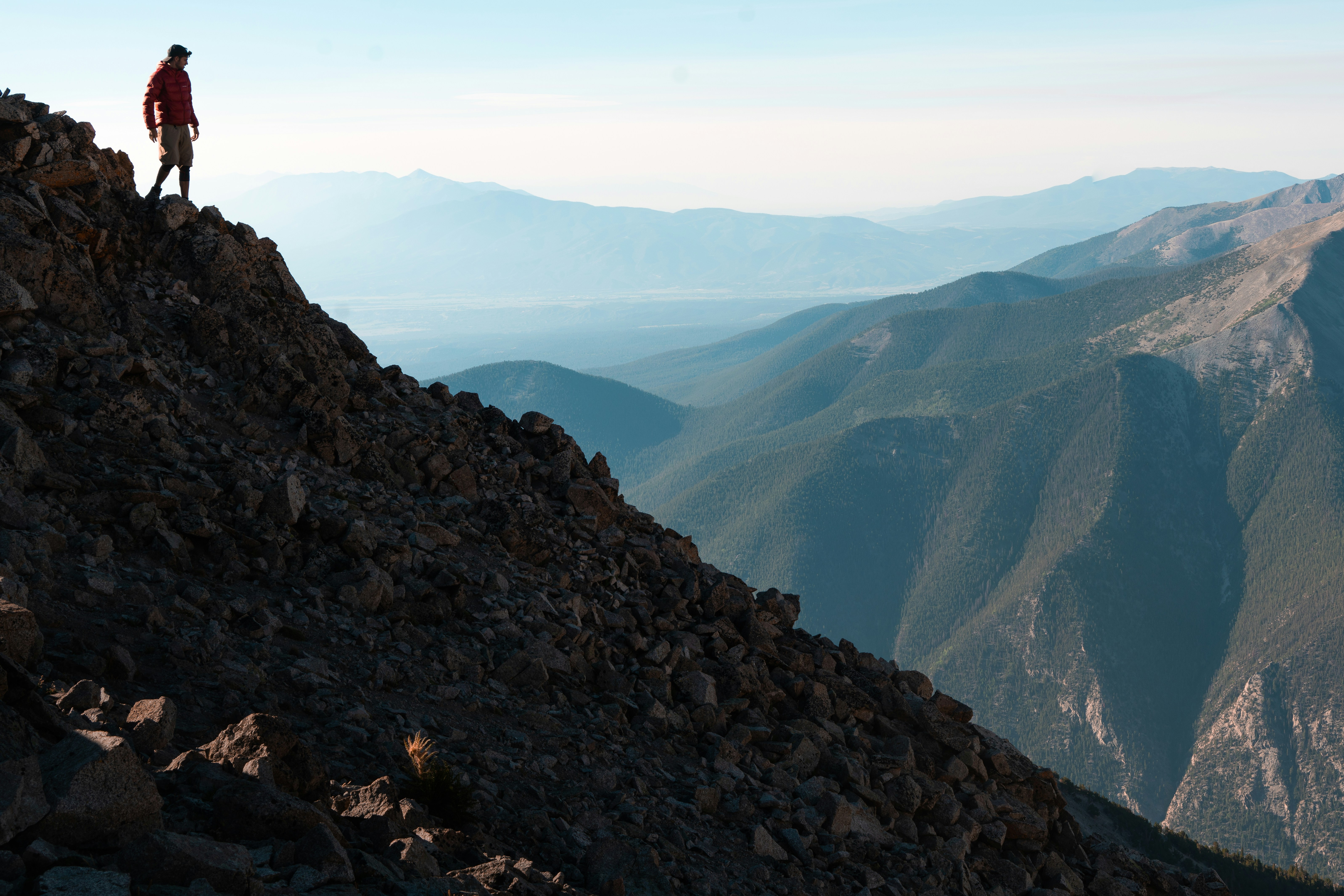 Silhouetted hiker on a rocky mountain ridge with expansive forested peaks under a clear sky.