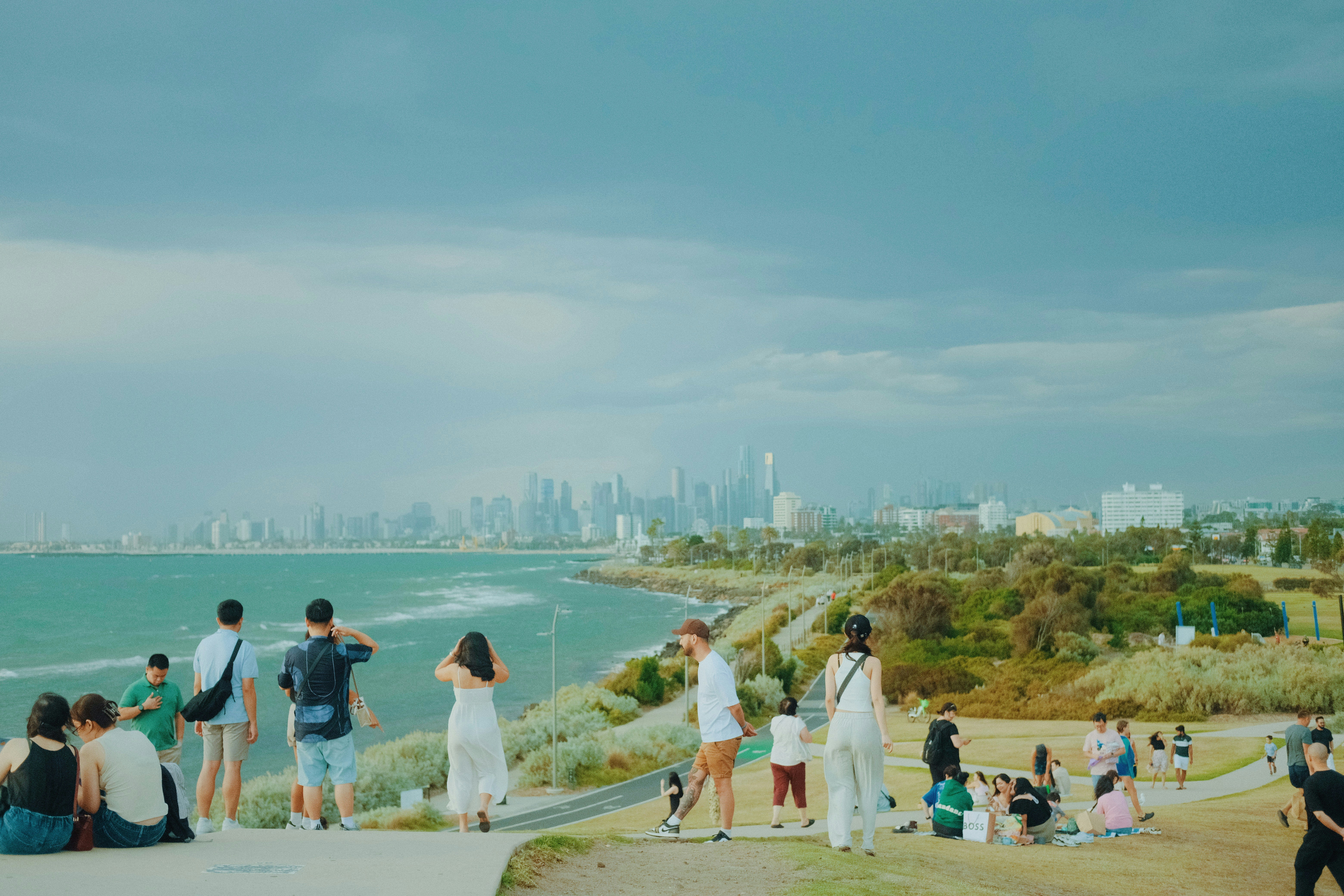 People gather along a coastal pathway with a sprawling city skyline in the distance under a vast blue sky.