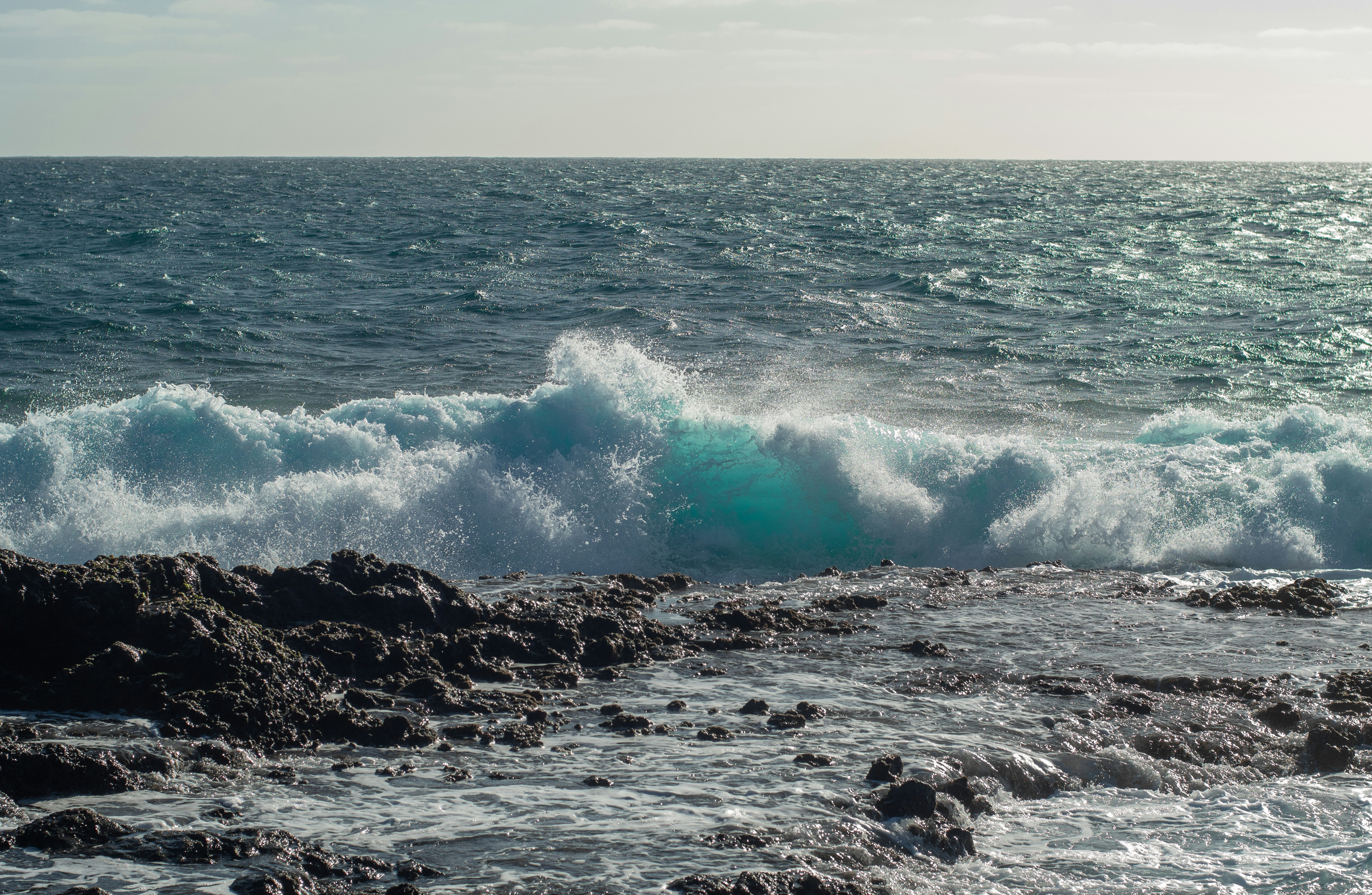 Ocean waves crash energetically against dark, rugged rocks with vibrant turquoise hues under soft lighting.