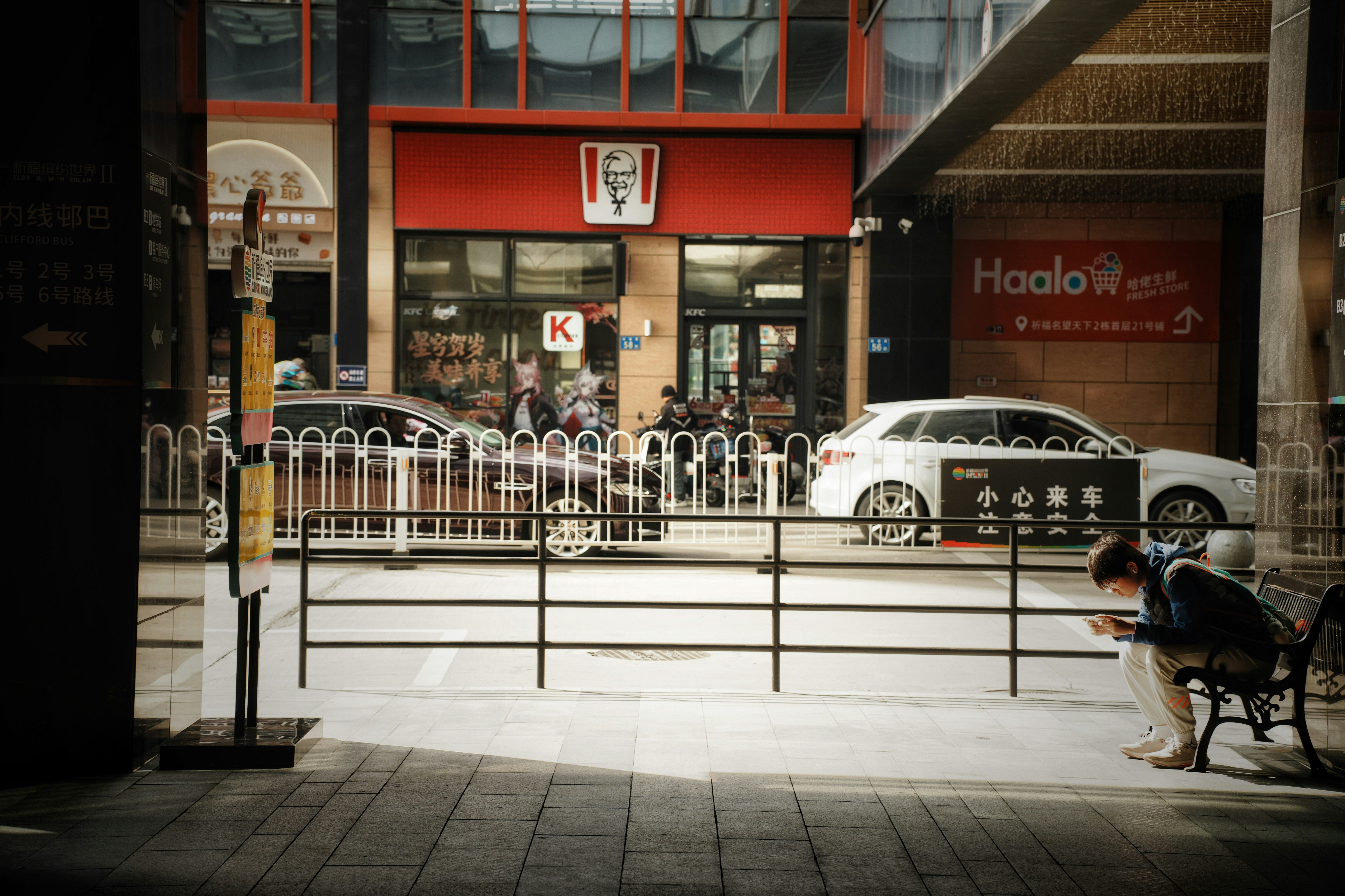 Person seated on a bench engrossed in a phone, with bustling city streets and colorful storefronts in the background.