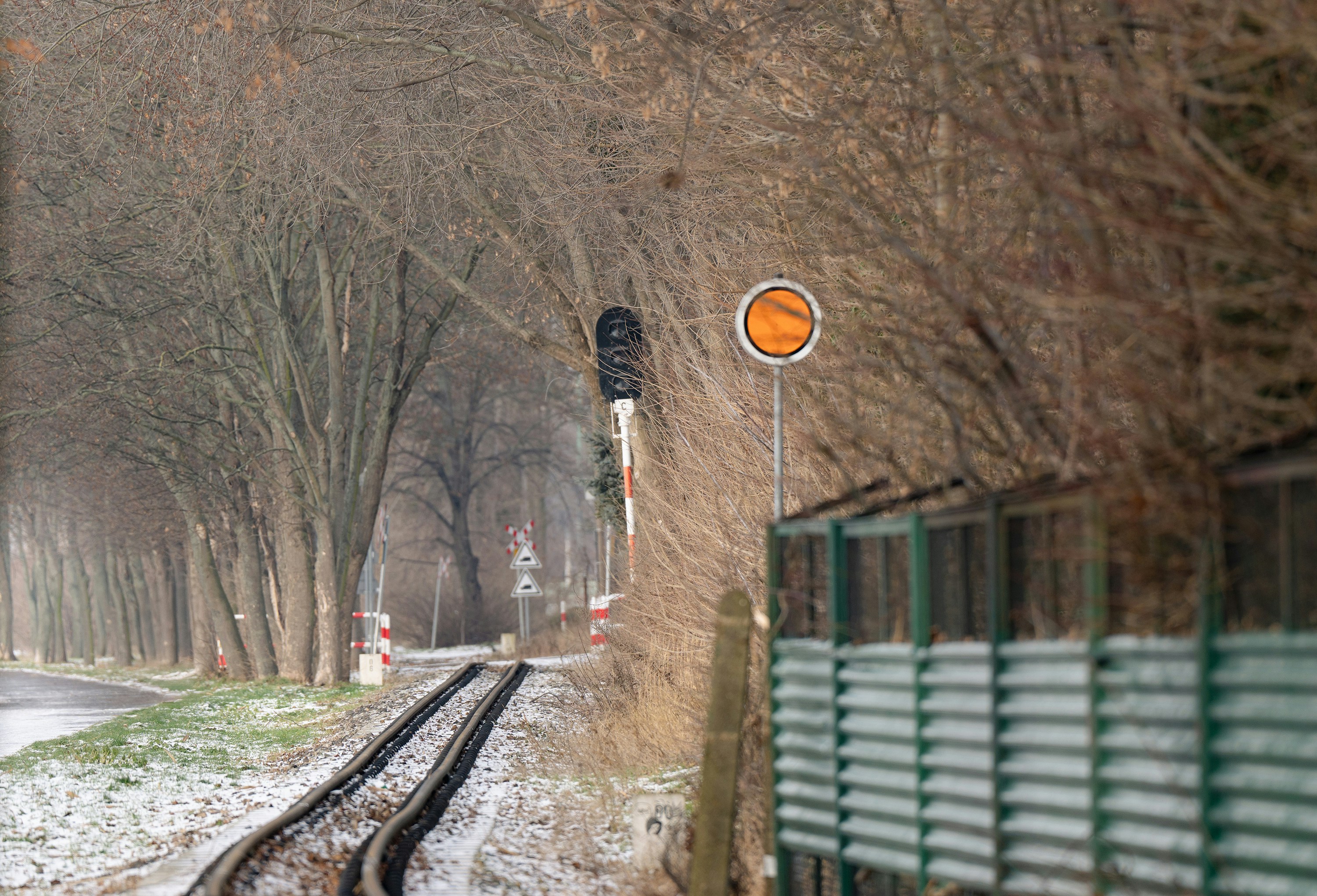 Snow-dusted railway curves through a leafless forest, accented by an orange sign and a rustic fence.