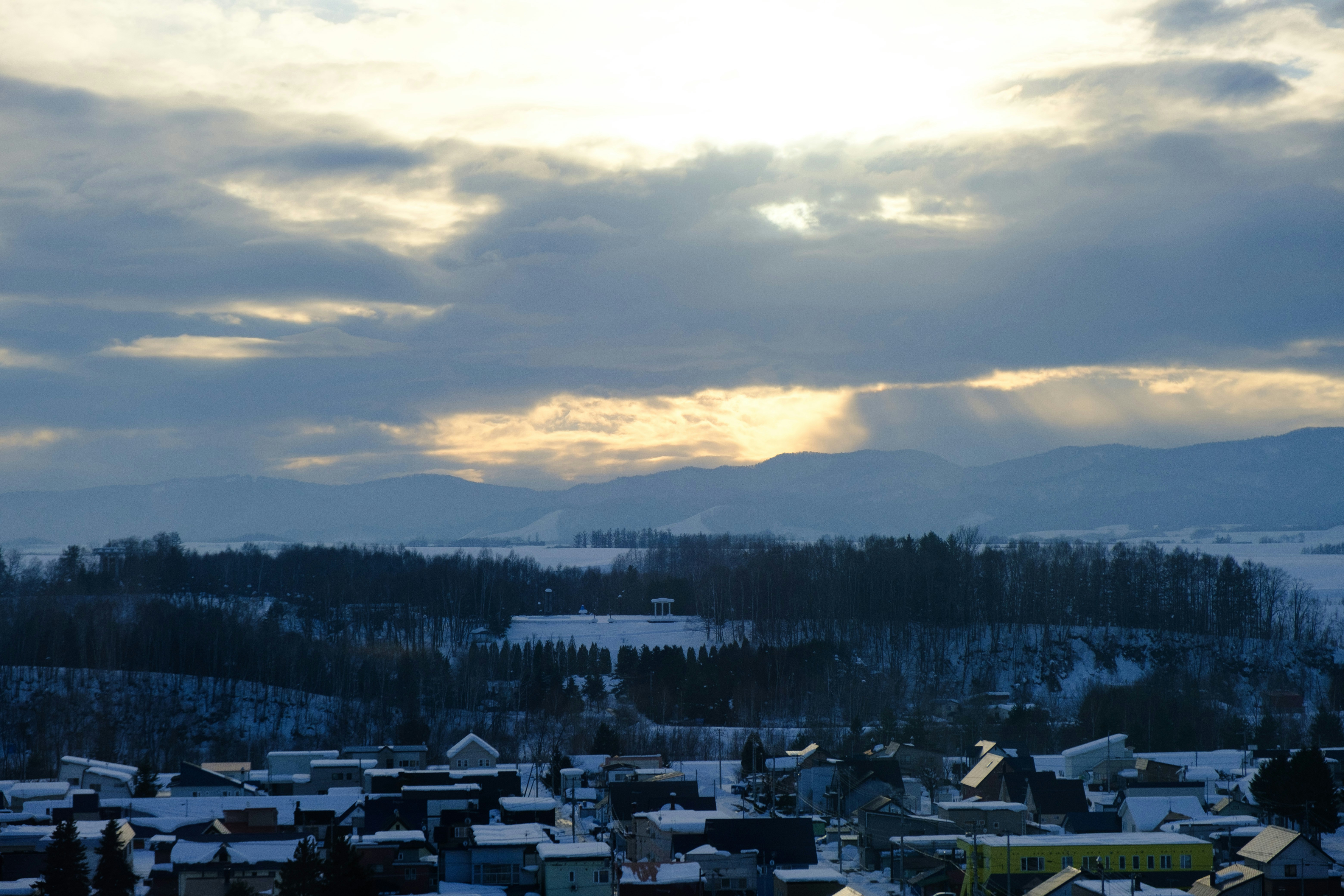 Sunrise casts a warm glow over a snow-covered town and distant mountains under a cloudy sky.