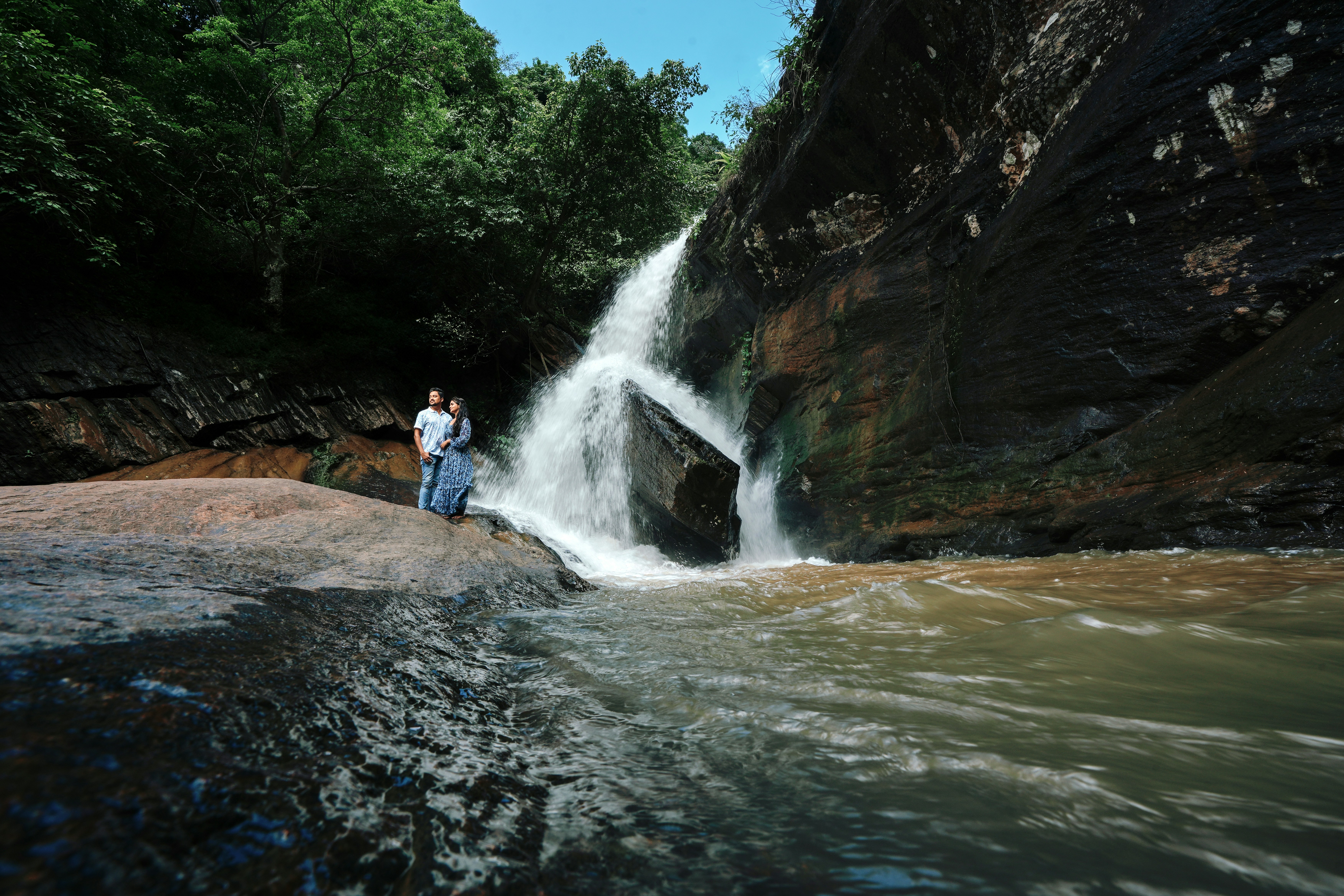 Couple stands beside a waterfall amid lush greenery and rugged rocks, creating a tranquil atmosphere.