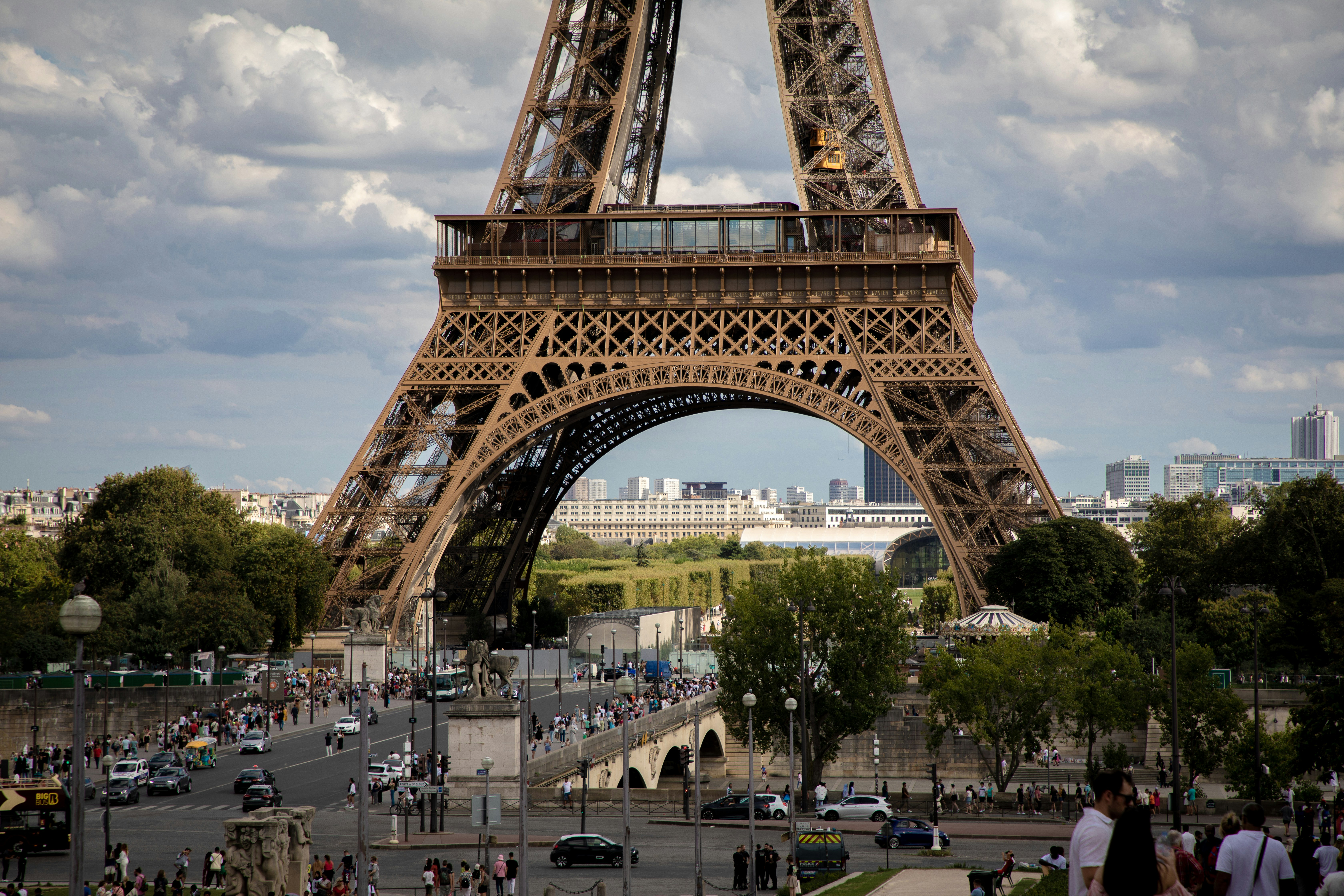 Eiffel Tower's intricate ironwork rises above a lively Paris street with lush greenery and scattered city life under a cloudy sky.