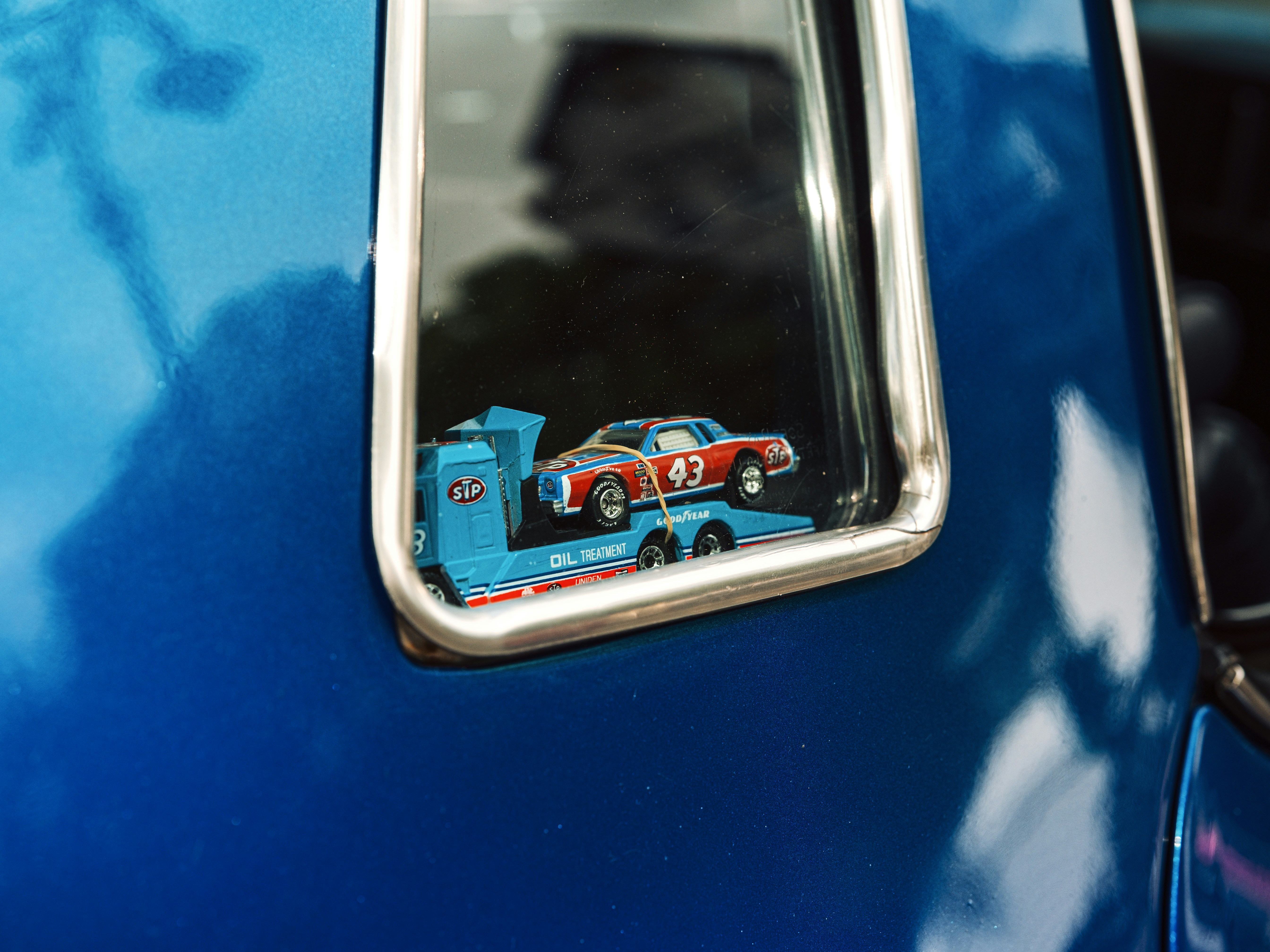 Miniature vintage race car reflected in a vibrant blue vehicle window, framed by chrome trim.