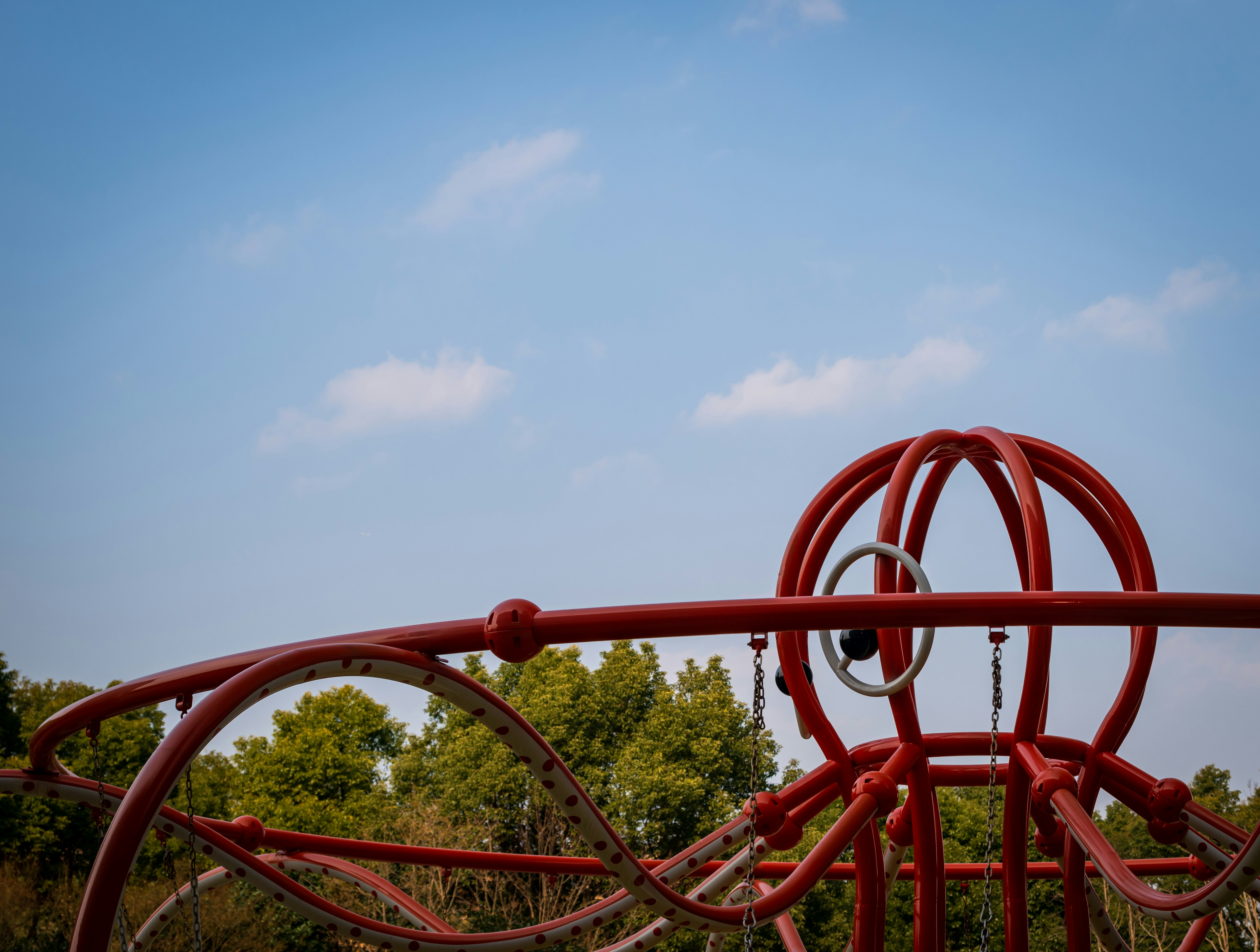 Bright red playground structure resembling an octopus against a clear blue sky, surrounded by lush greenery.