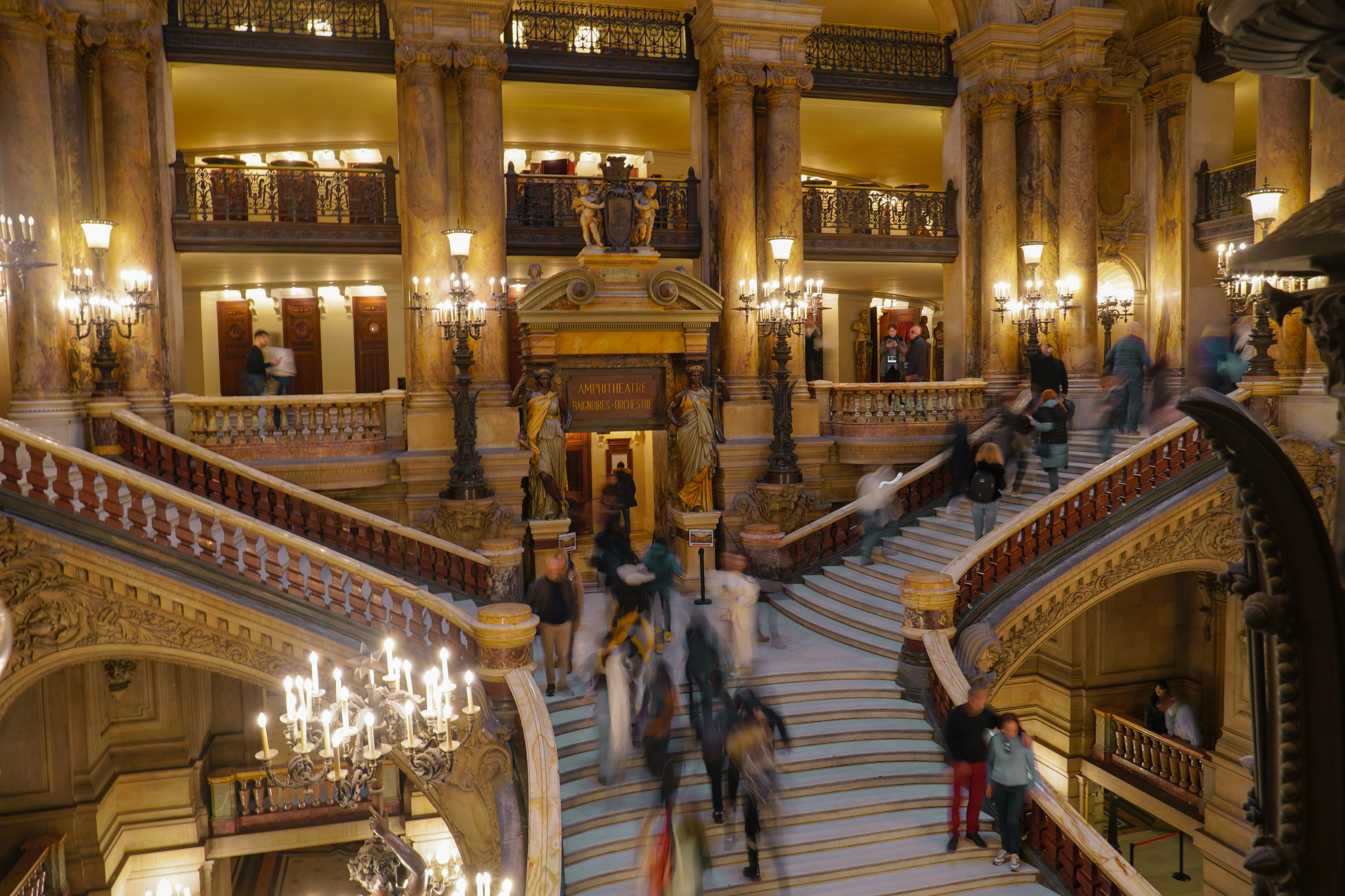 Blurred motion of visitors ascending the ornate staircase in a lavishly lit opera house.