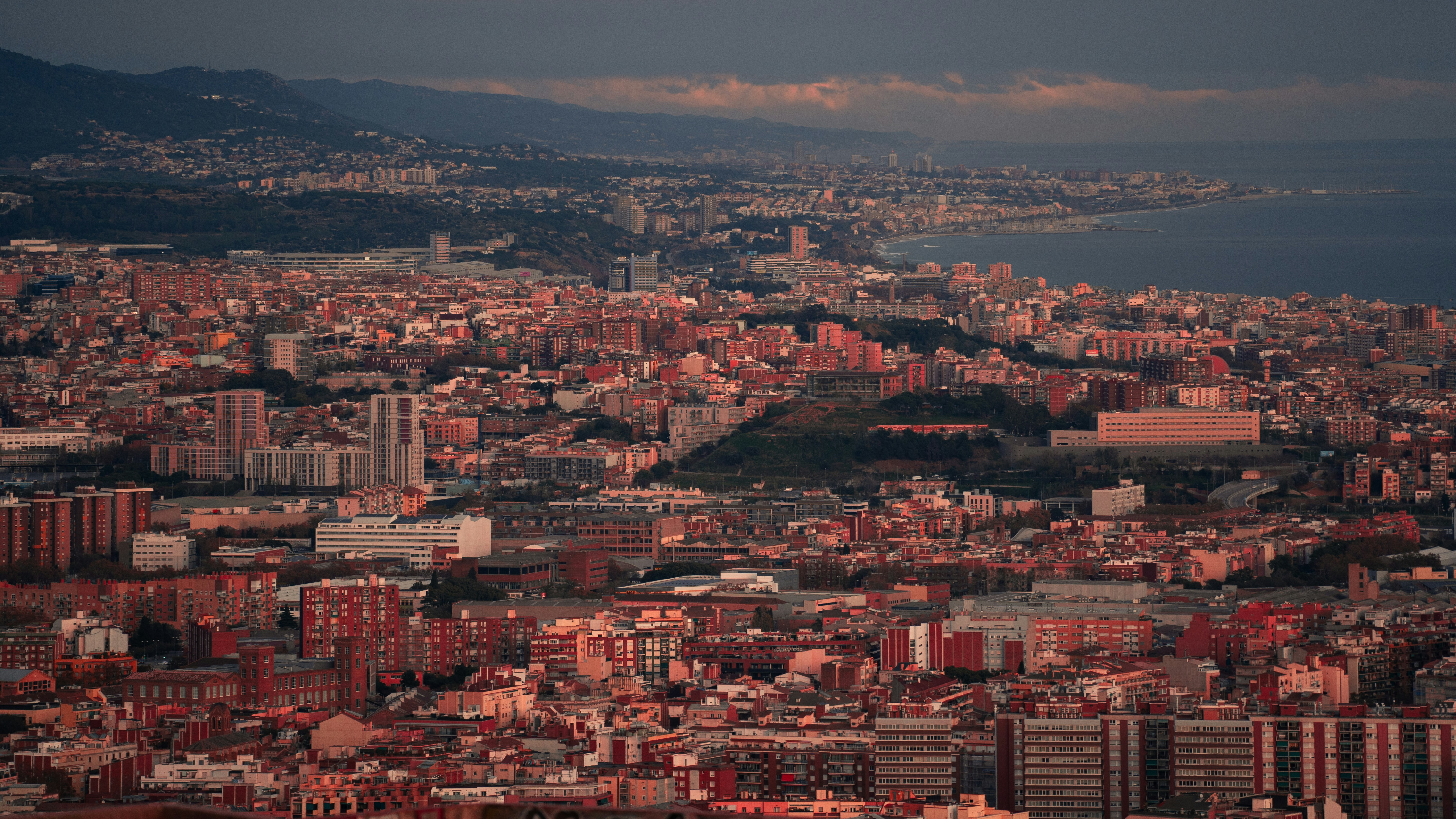 Urban sprawl with red rooftops under warm evening light, bordered by the sea and shadowed hills.