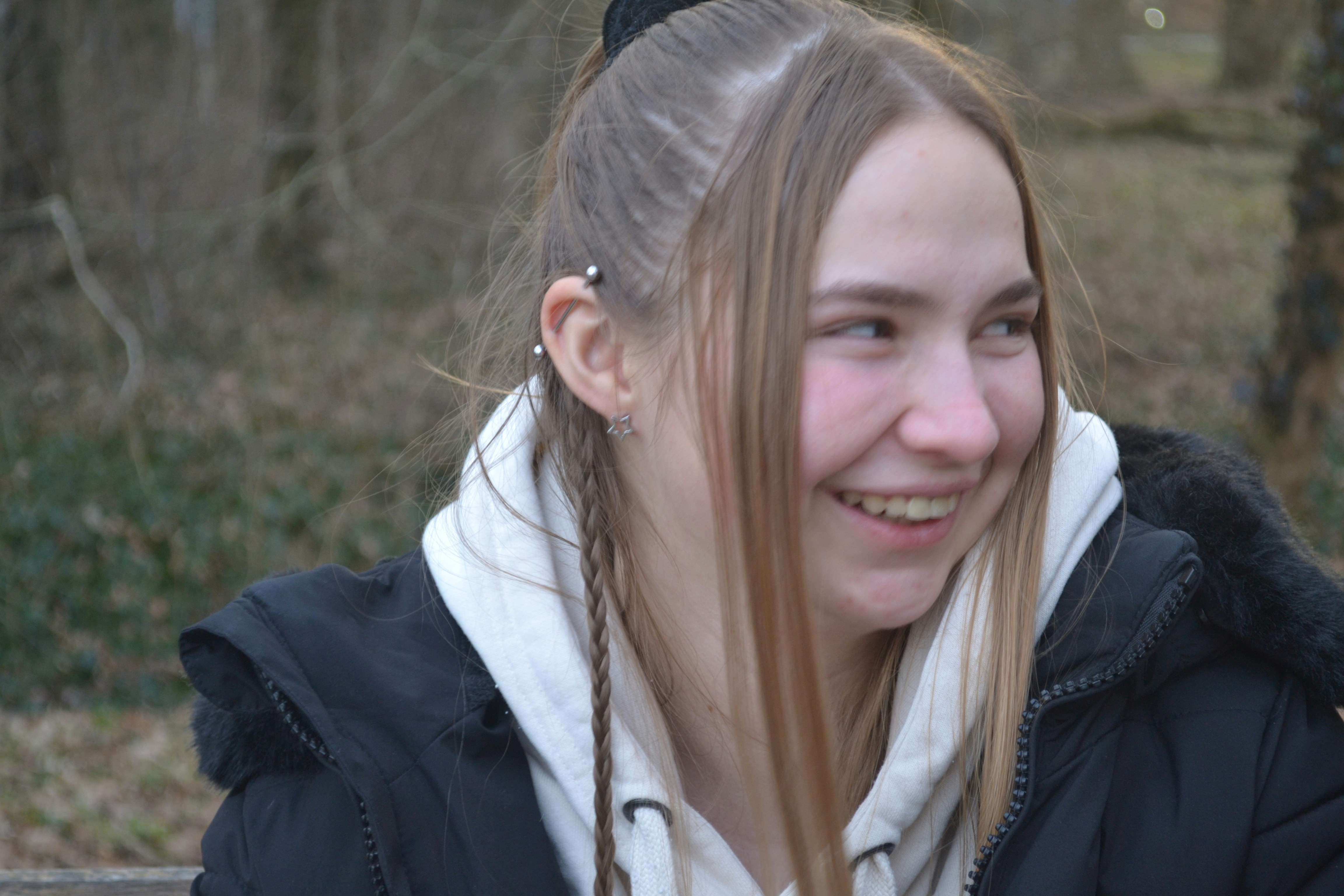 Young woman laughing in a forest with light brown hair and black jacket against a blurred woodland background.