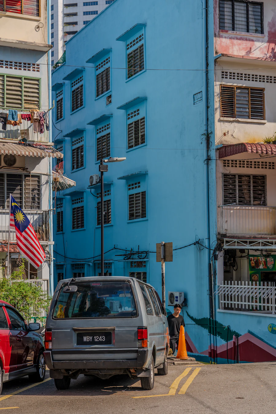 Bright blue apartment facade framed by older buildings on a tight urban street, with parked vehicles, a Malaysian flag, and a pedestrian near the curb.