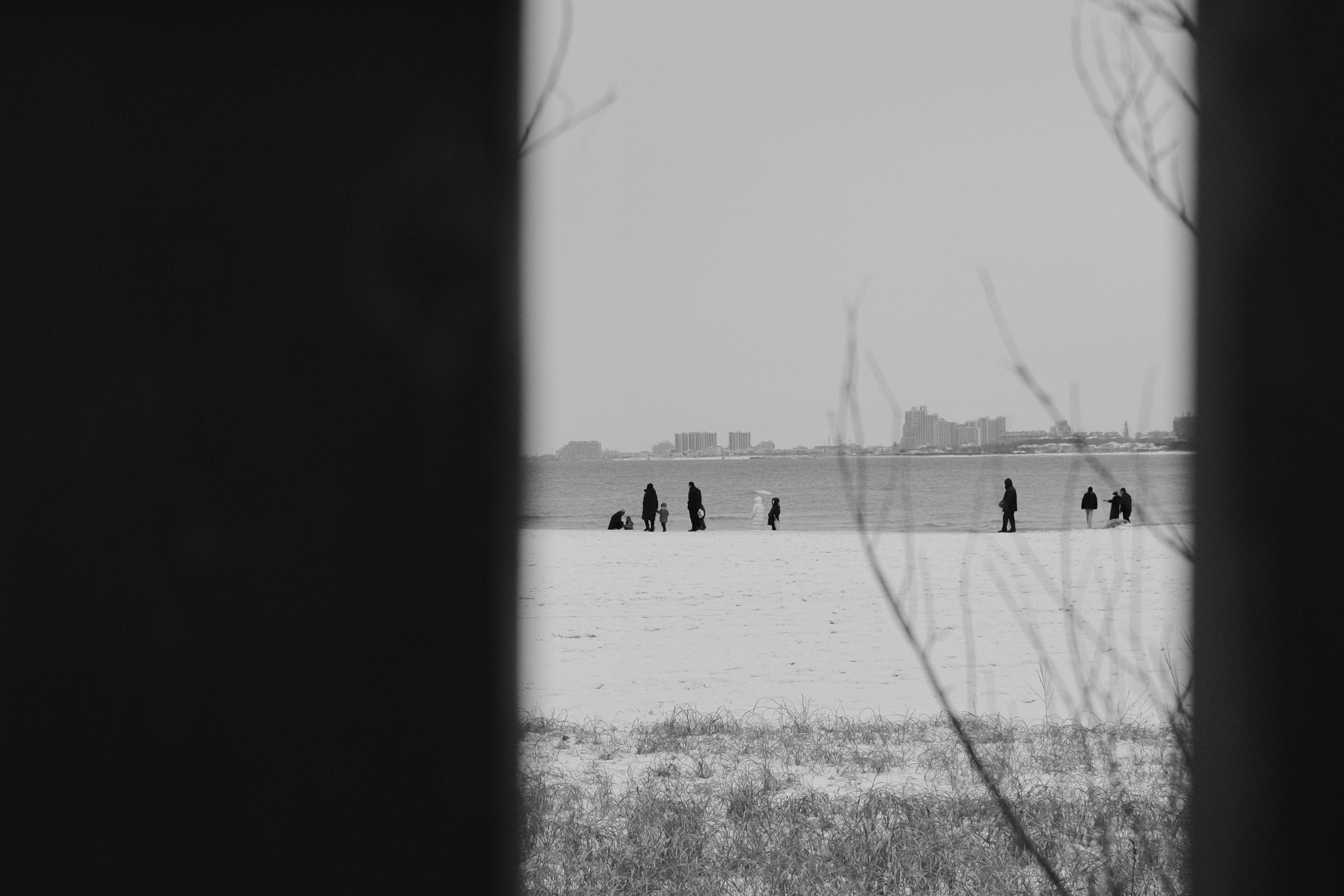 People walking on a snow-covered beach by a calm sea, framed by dark vertical elements and distant skyscrapers on the horizon.
