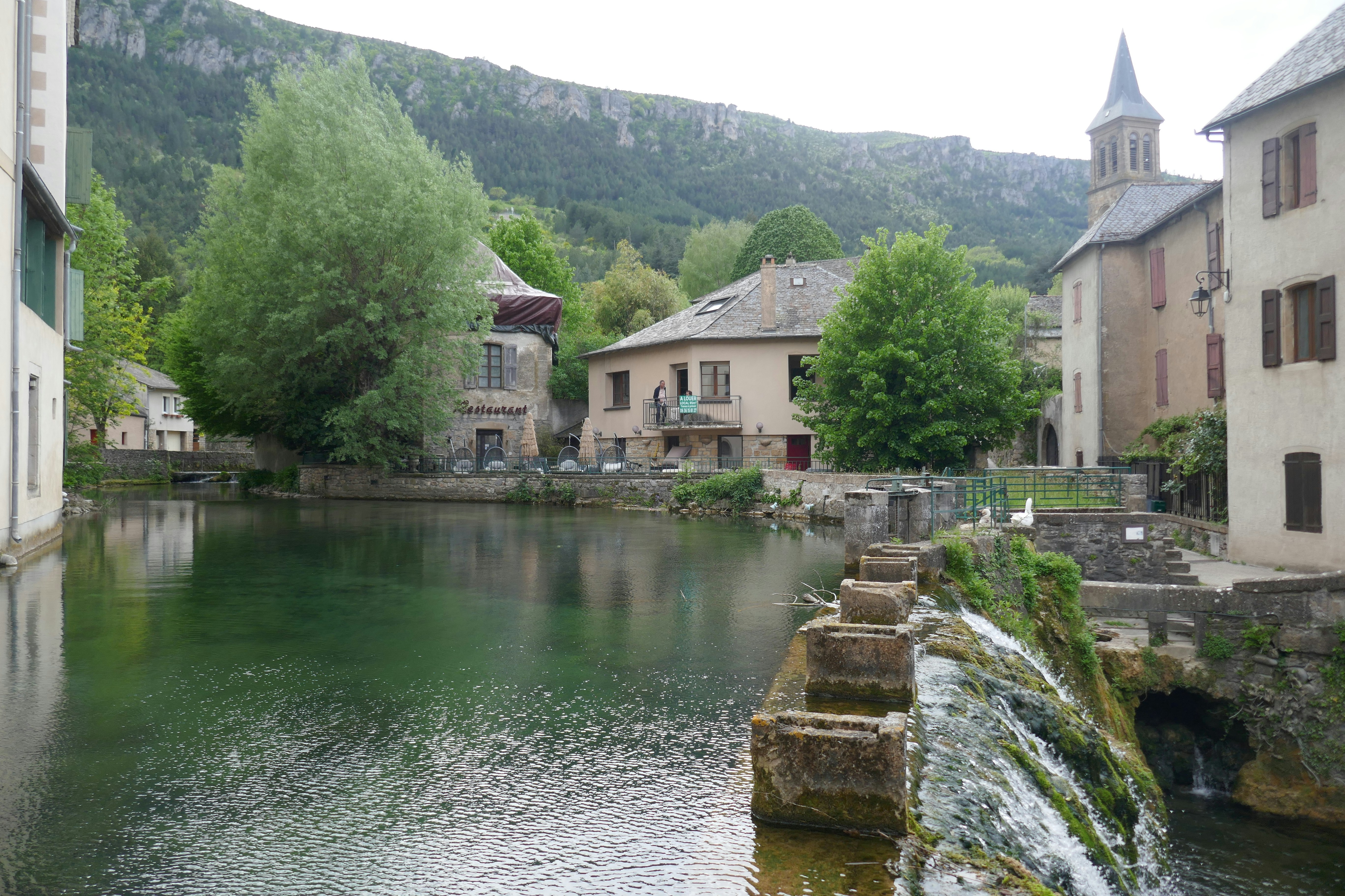 Charming village buildings reflected in a serene river with a waterfall in the foreground.