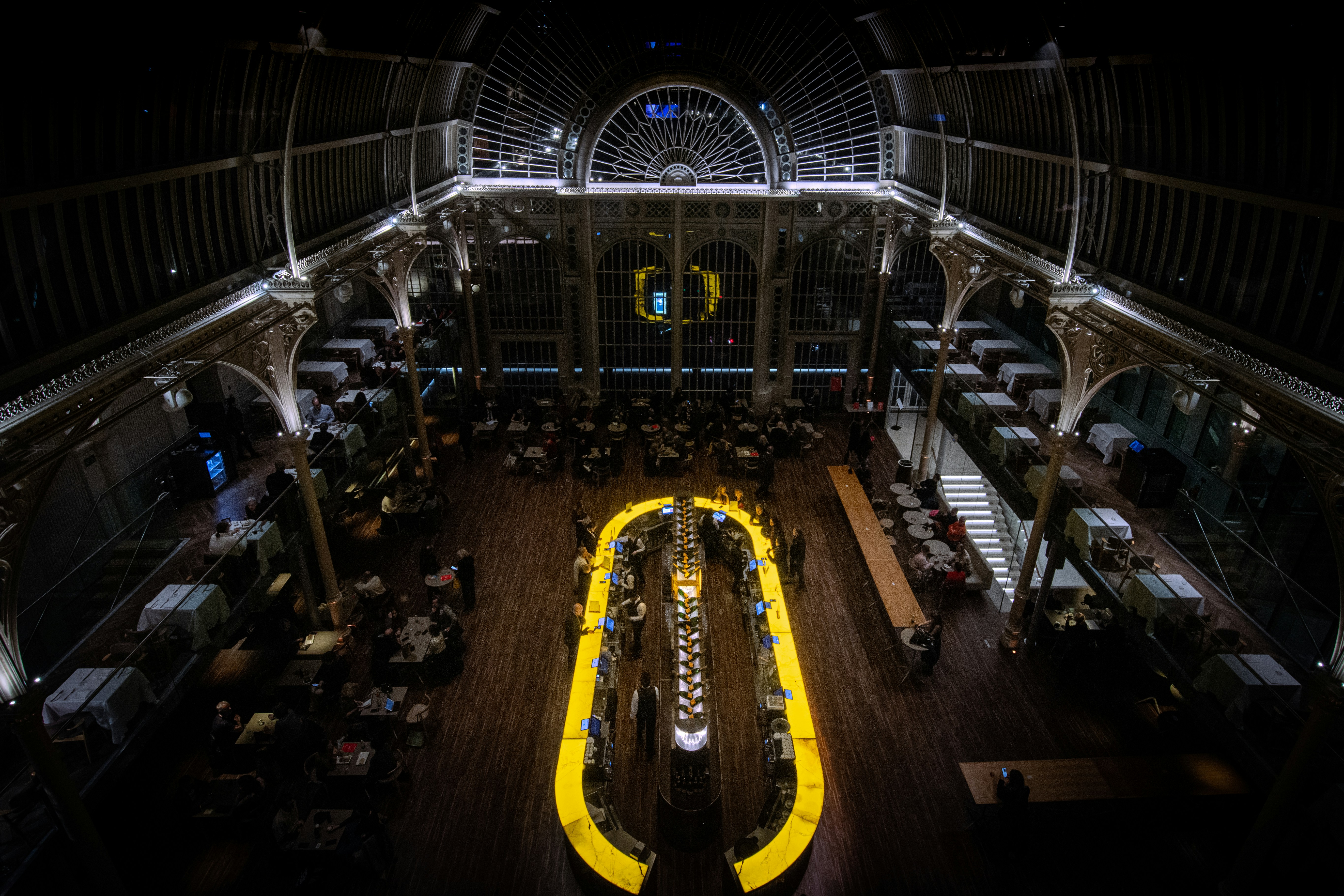 Dimly lit grand hall with a glowing oval bar at its center, under an ornate arched ceiling.