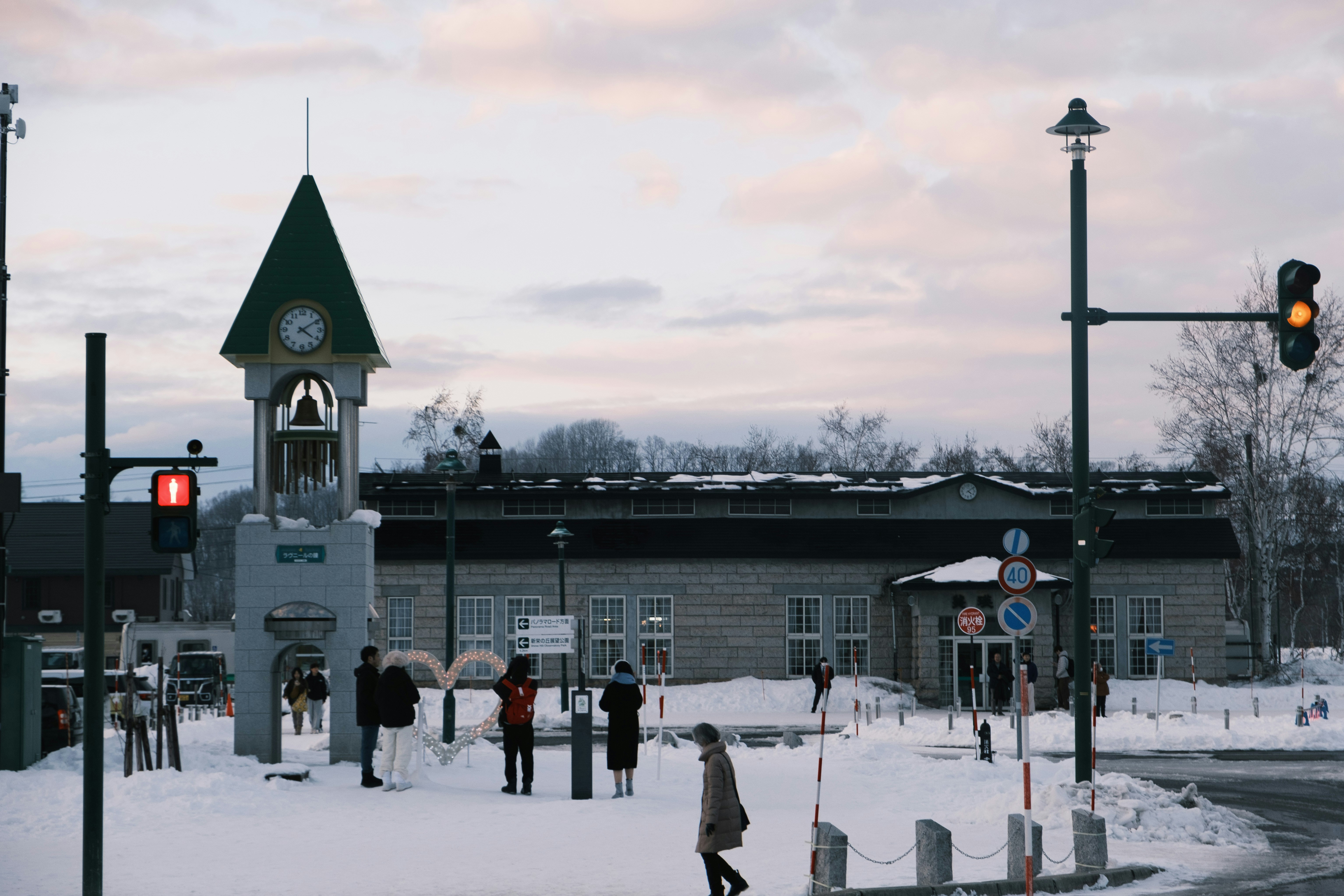 Snowy street scene with a clock tower and people walking under pastel evening sky.