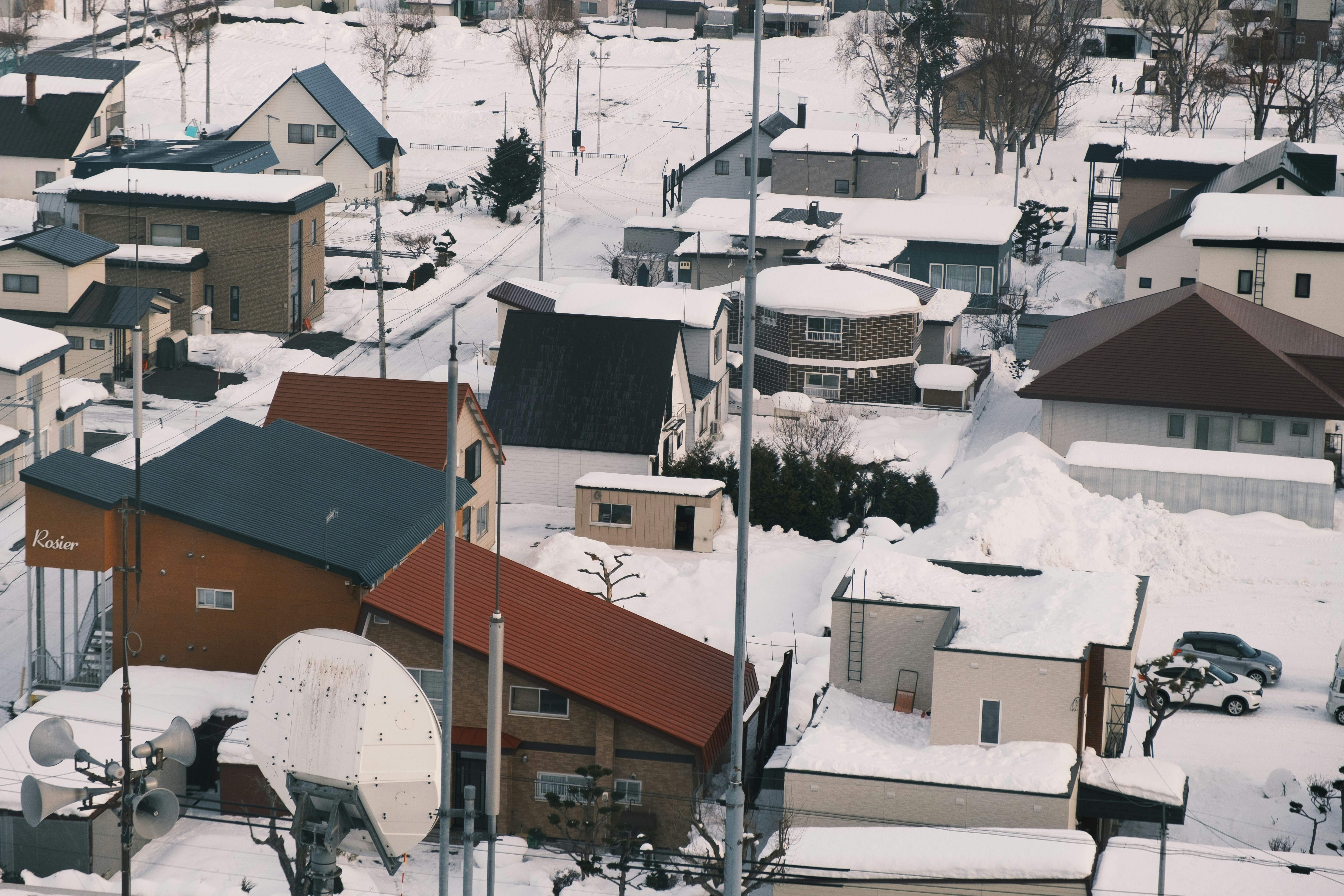 Snow-covered suburban neighborhood with modern and traditional homes, rooftops in muted colors contrasting against white snow.