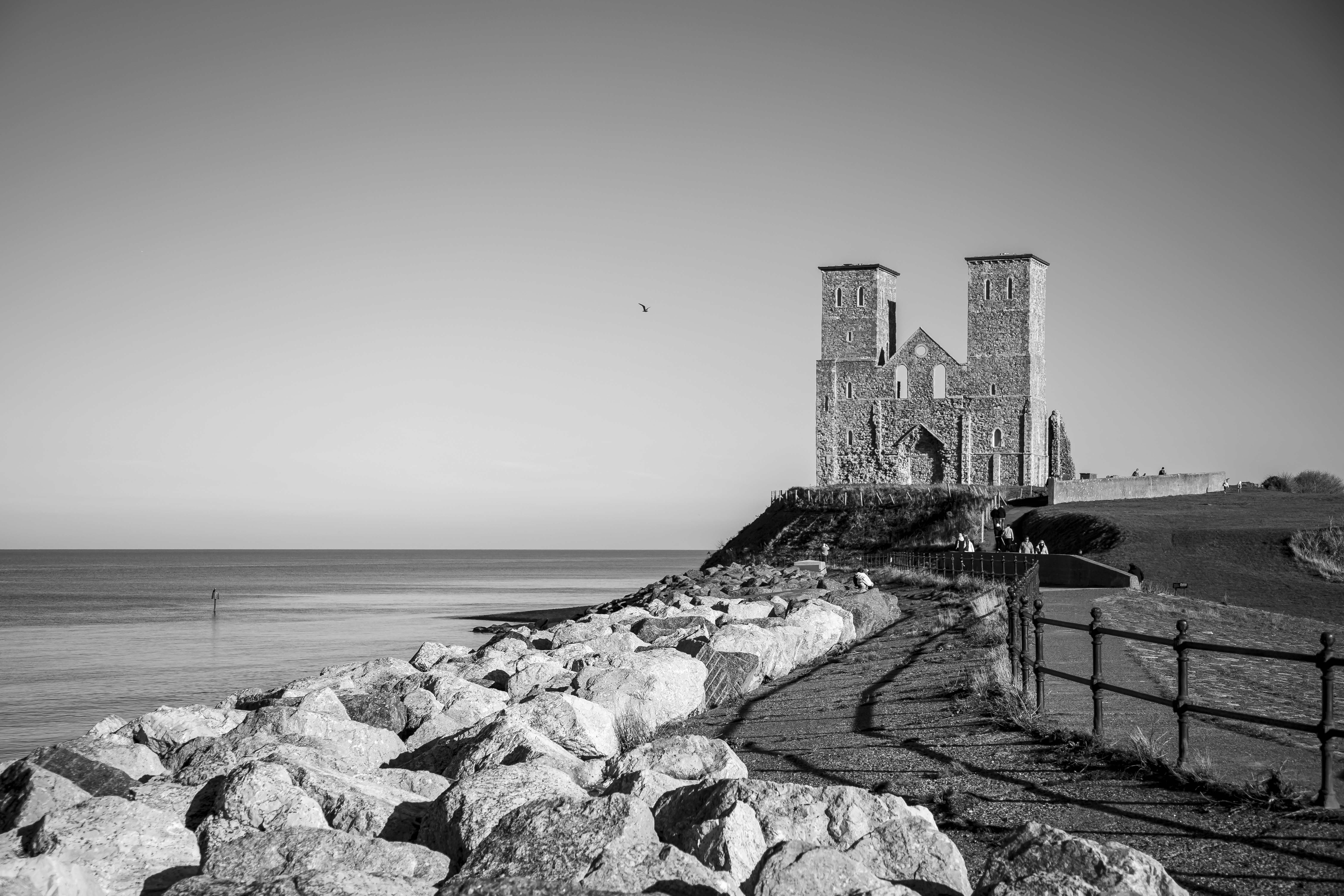Black and white photo of Reculver Towers with a seagull in flight against a serene coastal backdrop.