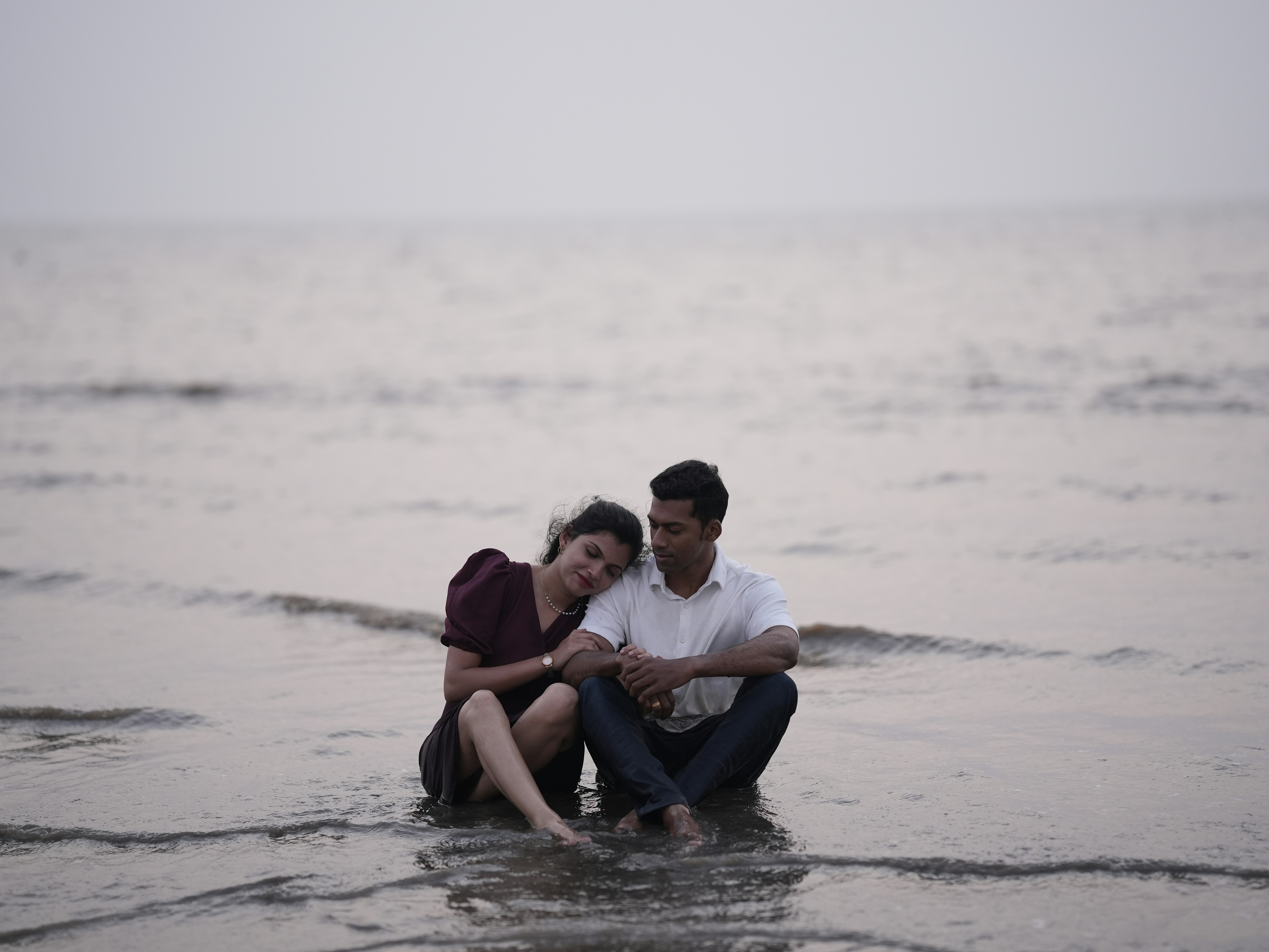 Couple sitting closely on a rock surrounded by gentle ocean waves under a muted sky.