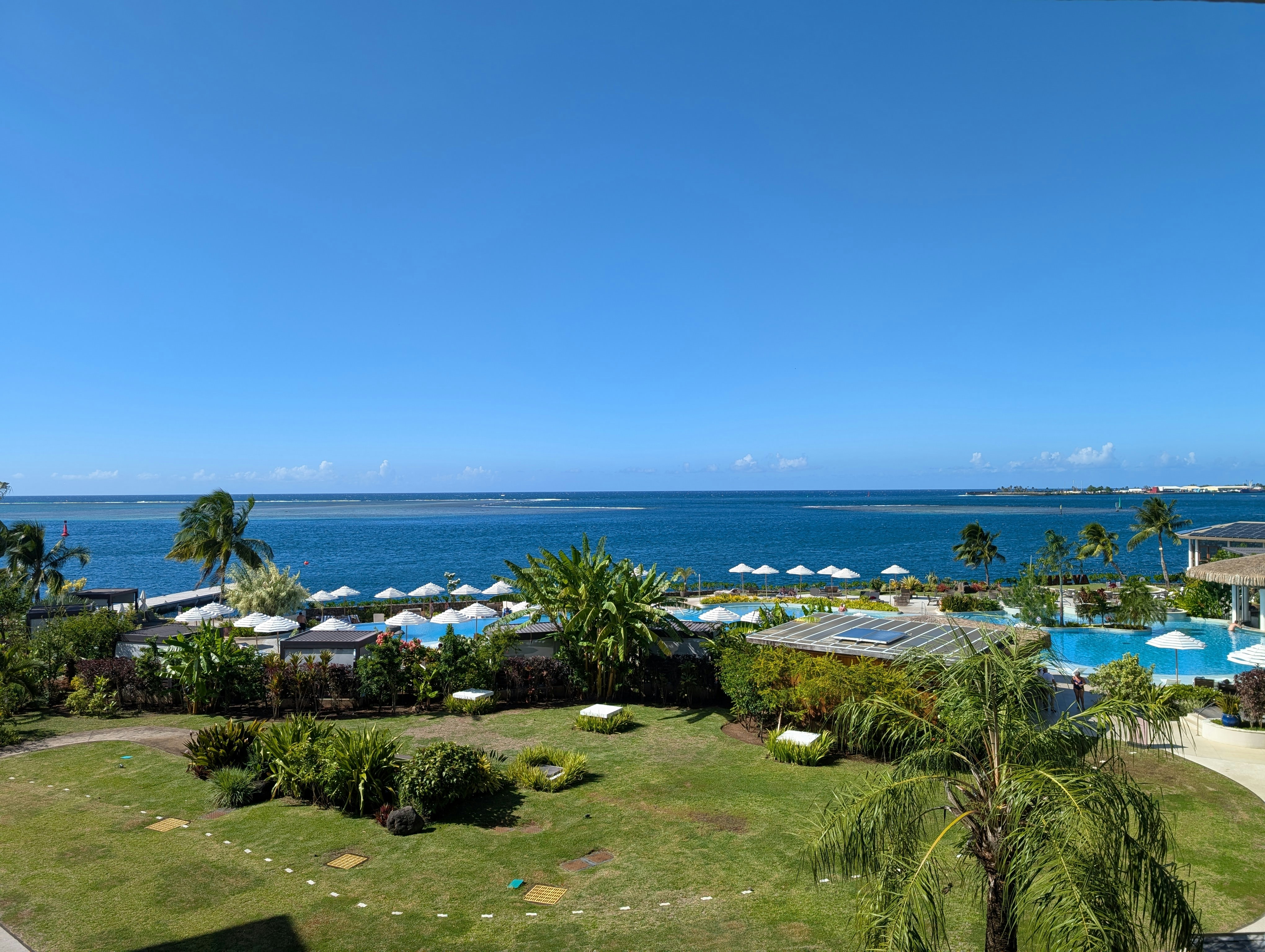 Coastal resort with palm trees, lush gardens, and distant ocean horizon under a clear blue sky.
