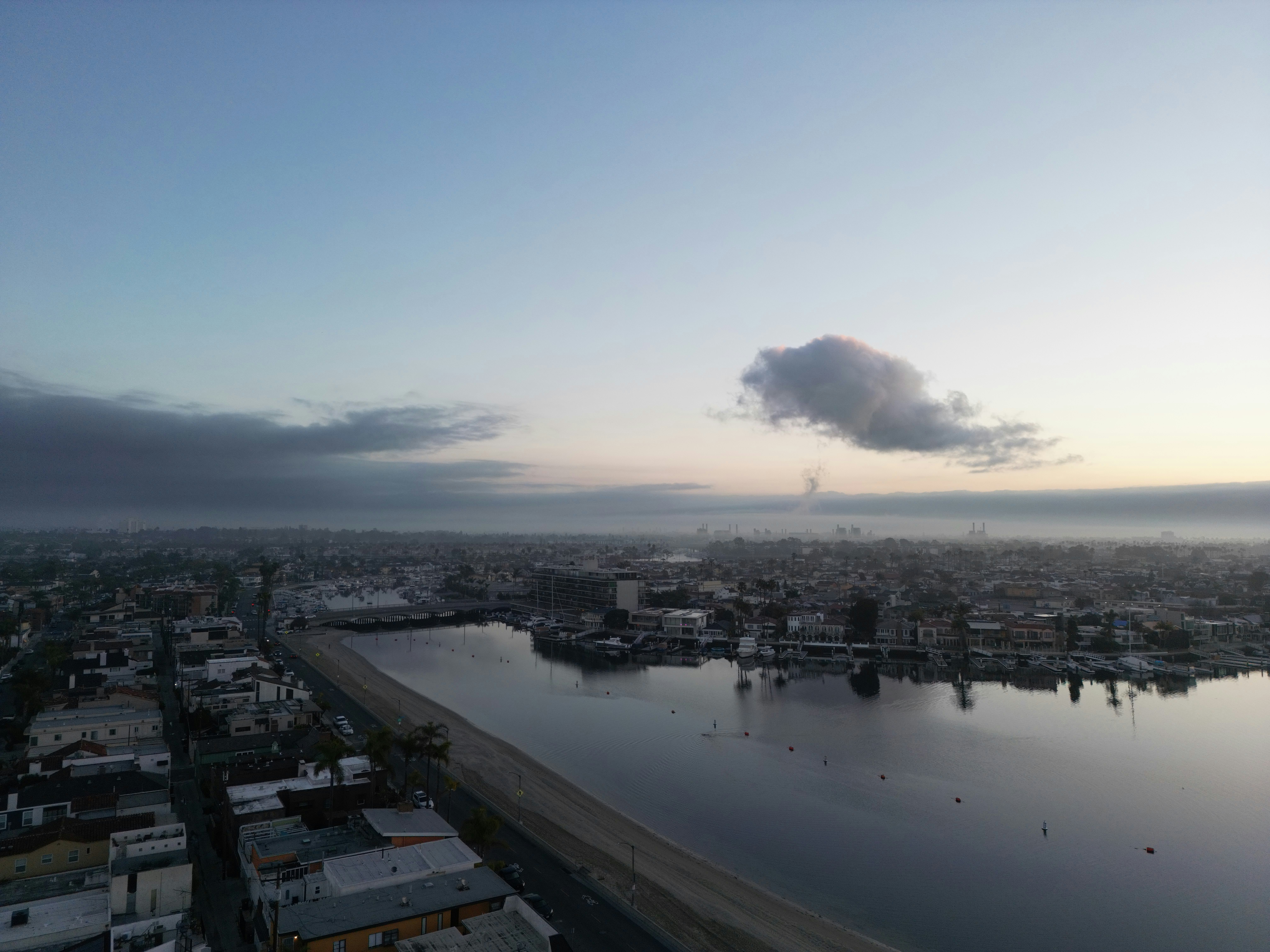 Coastal town at dawn with calm waters reflecting pastel sky hues and a solitary cloud enhancing depth.