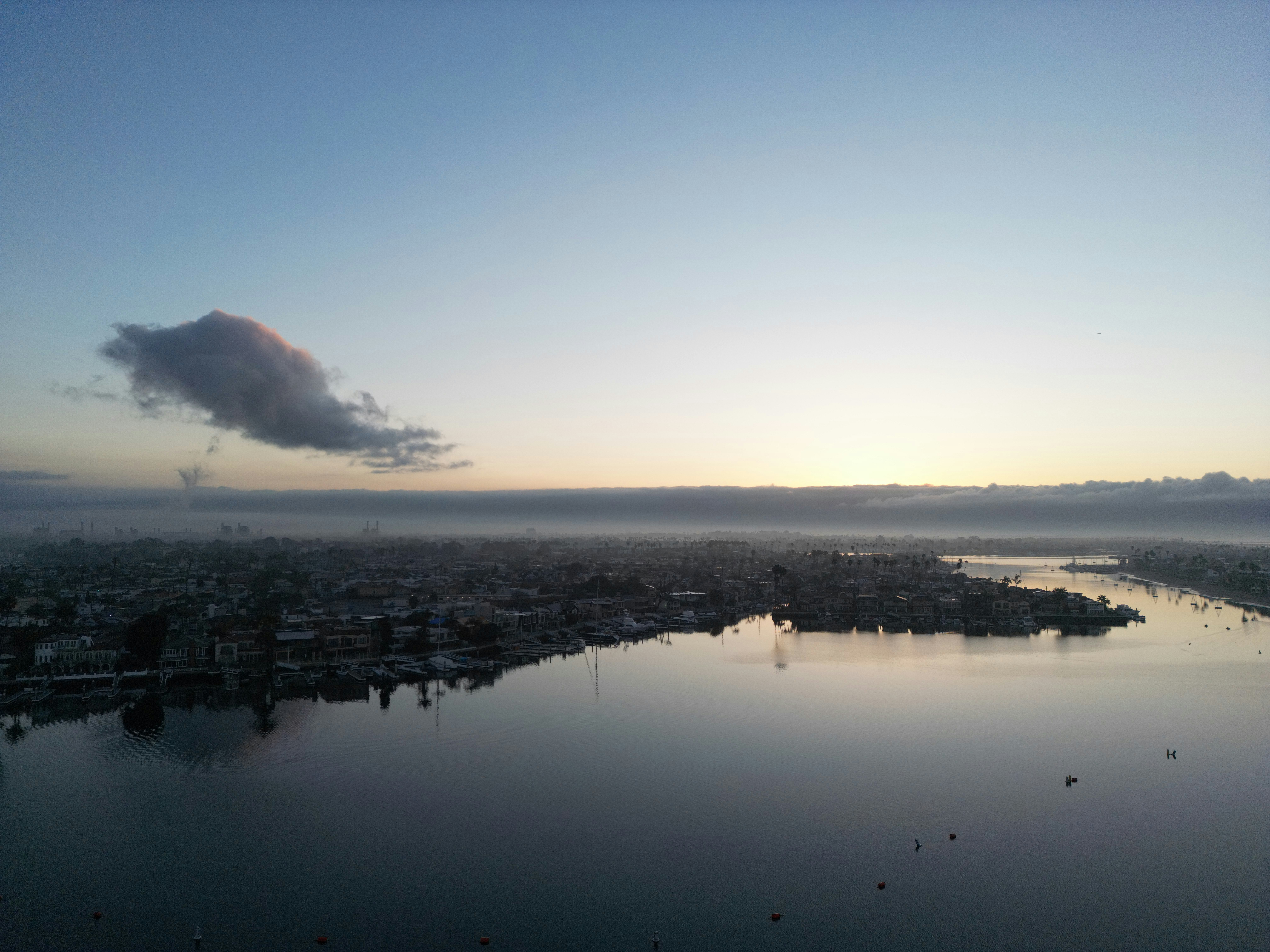 Aerial view of a peaceful coastal cityscape with calm waters reflecting the early morning sky and a solitary cloud above the horizon.