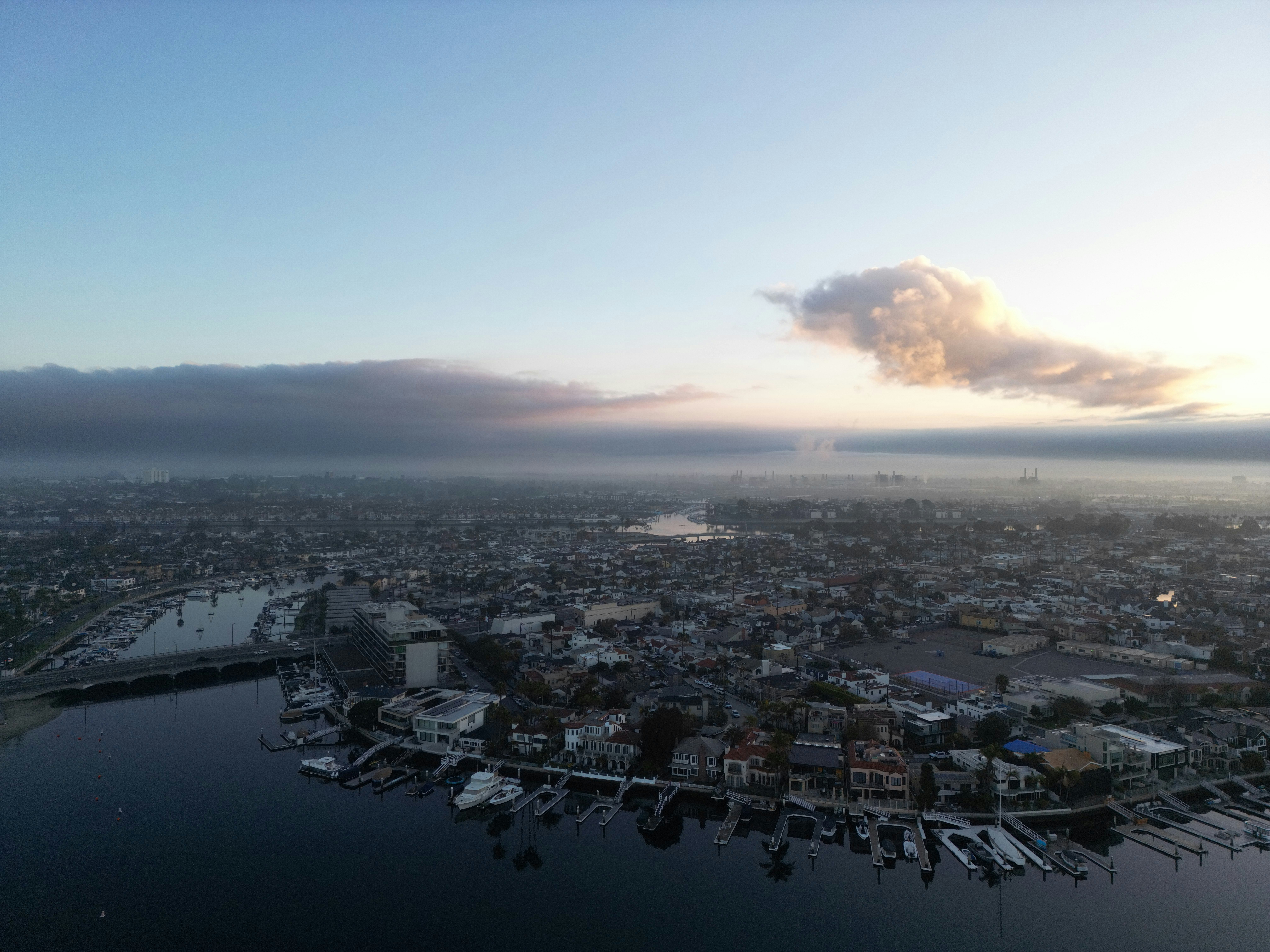 Coastal cityscape at dawn with calm waters reflecting early morning hues, featuring a marina and sprawling urban area beneath soft clouds.