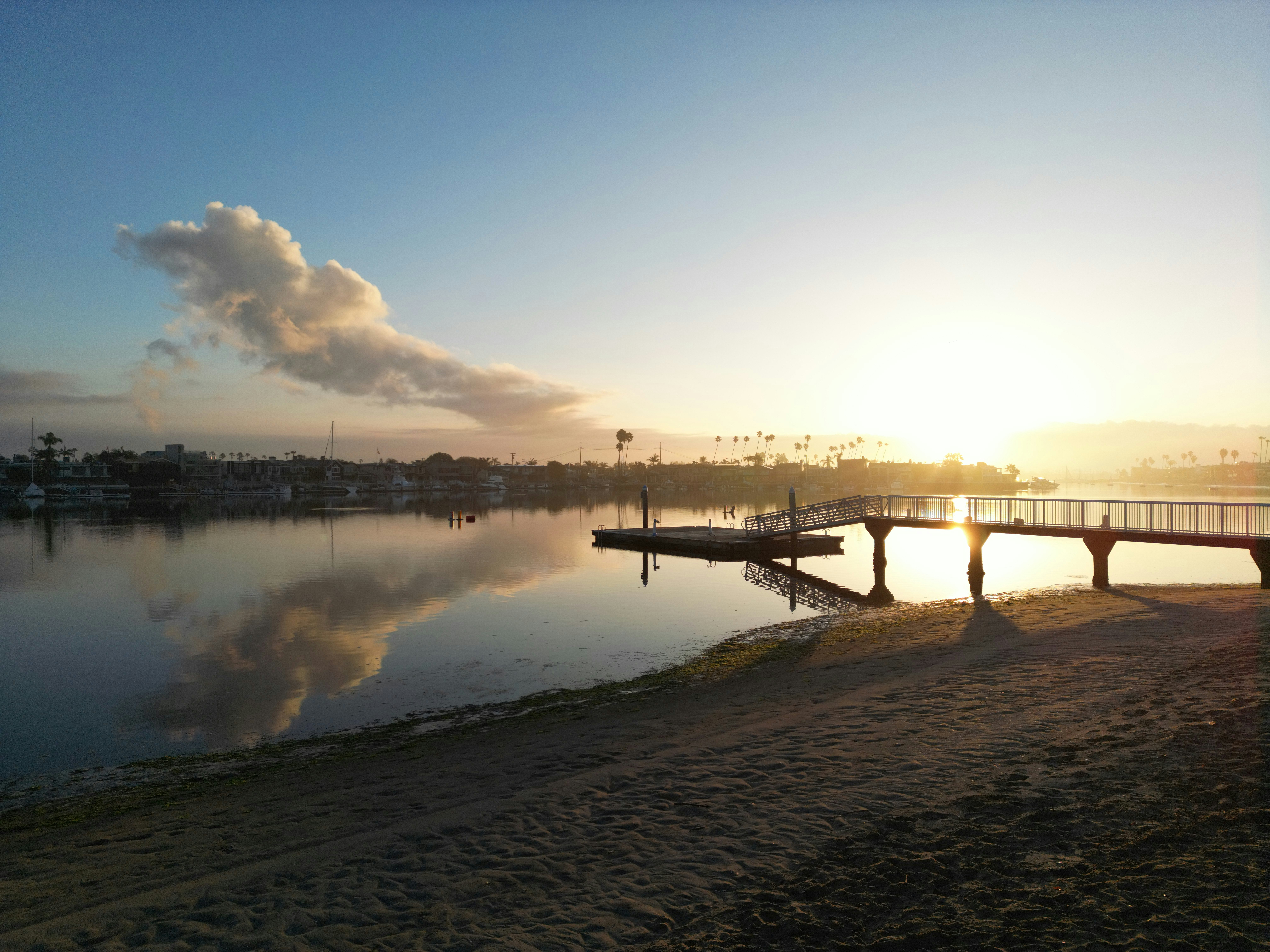 Silhouetted pier stretches over calm waters reflecting a cloud under a pastel sky at sunrise.