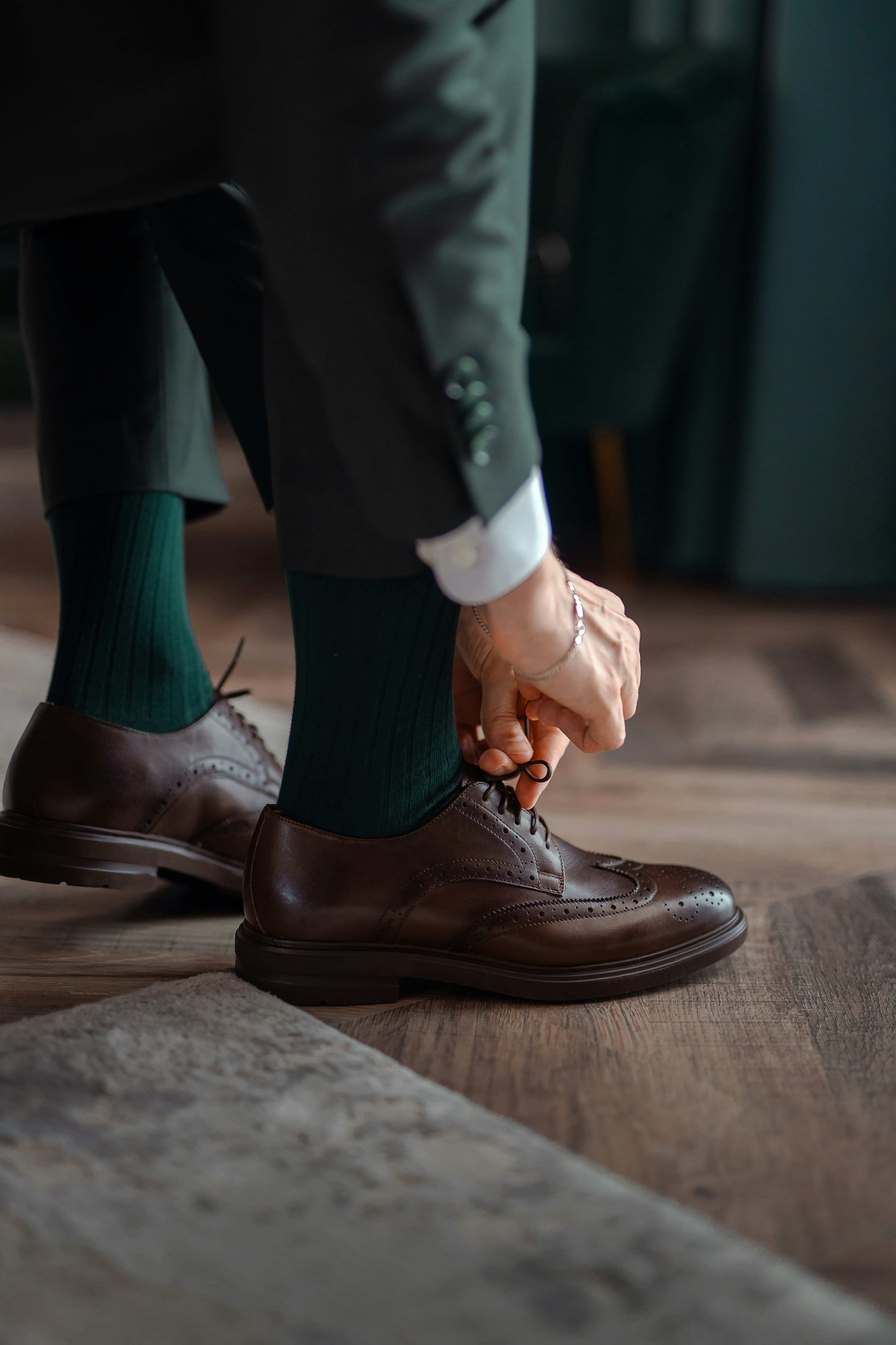 A person tying the laces of a brown leather dress shoe, wearing dark green socks and a dark suit, with a white shirt cuff visible. The wooden floor is in the background. | A man in a suit tying his shoes