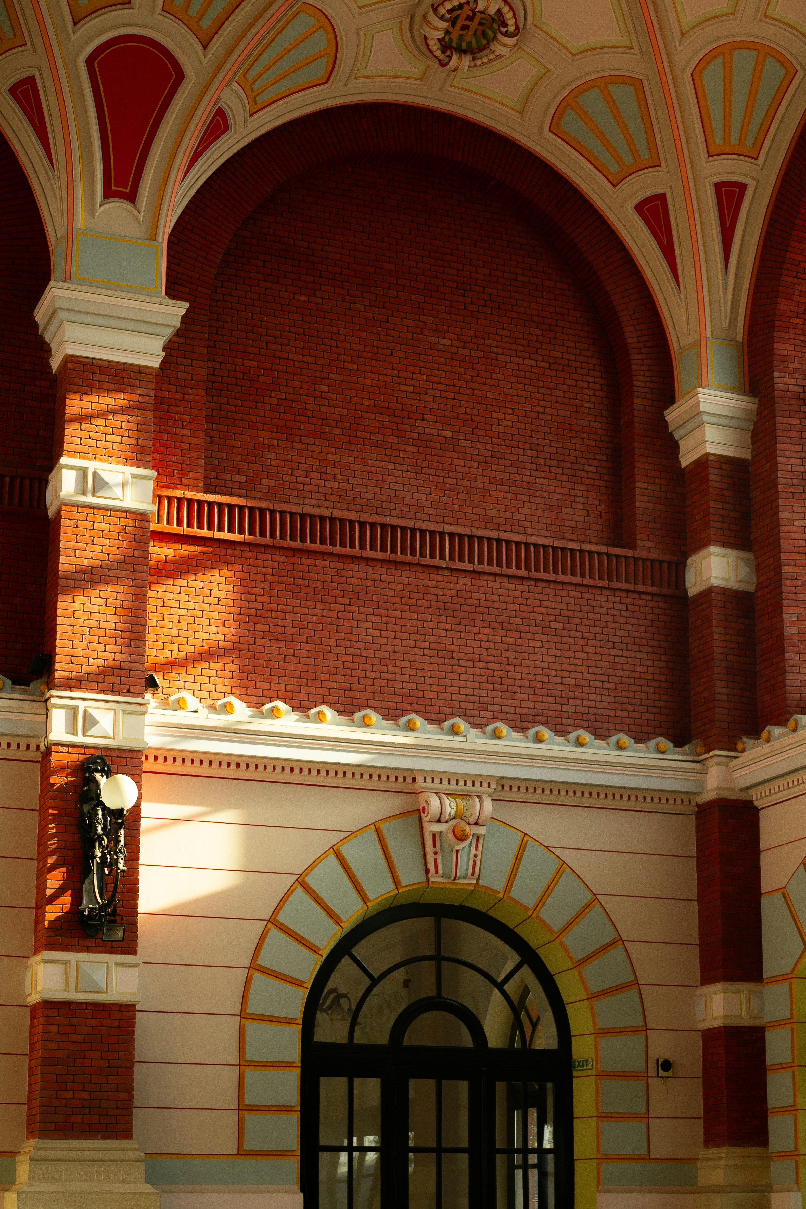 A beautifully ornate interior with a large arched brick wall framed by columns, featuring intricate detailing and a painted ceiling with geometric patterns. Below the arch is a lighter-colored section with a decorative molding and an arched doorway with a black frame and glass panels. The surrounding area is adorned with patterns and a decorative light fixture, suggesting a historical or significant building. | A building with a clock on the front of it
