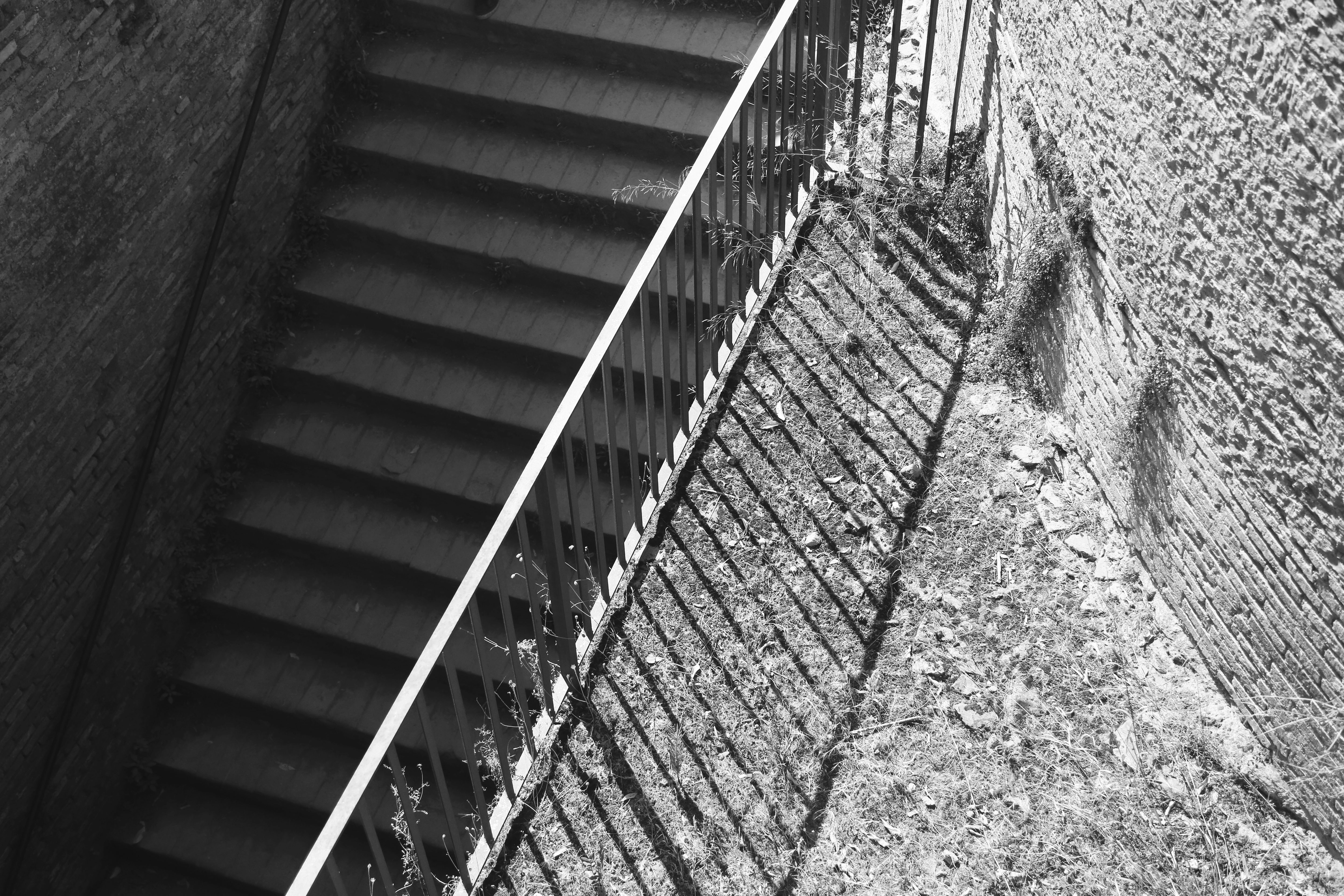 Monochrome staircase with sunlight casting intricate shadow patterns through the railing onto textured brick walls.