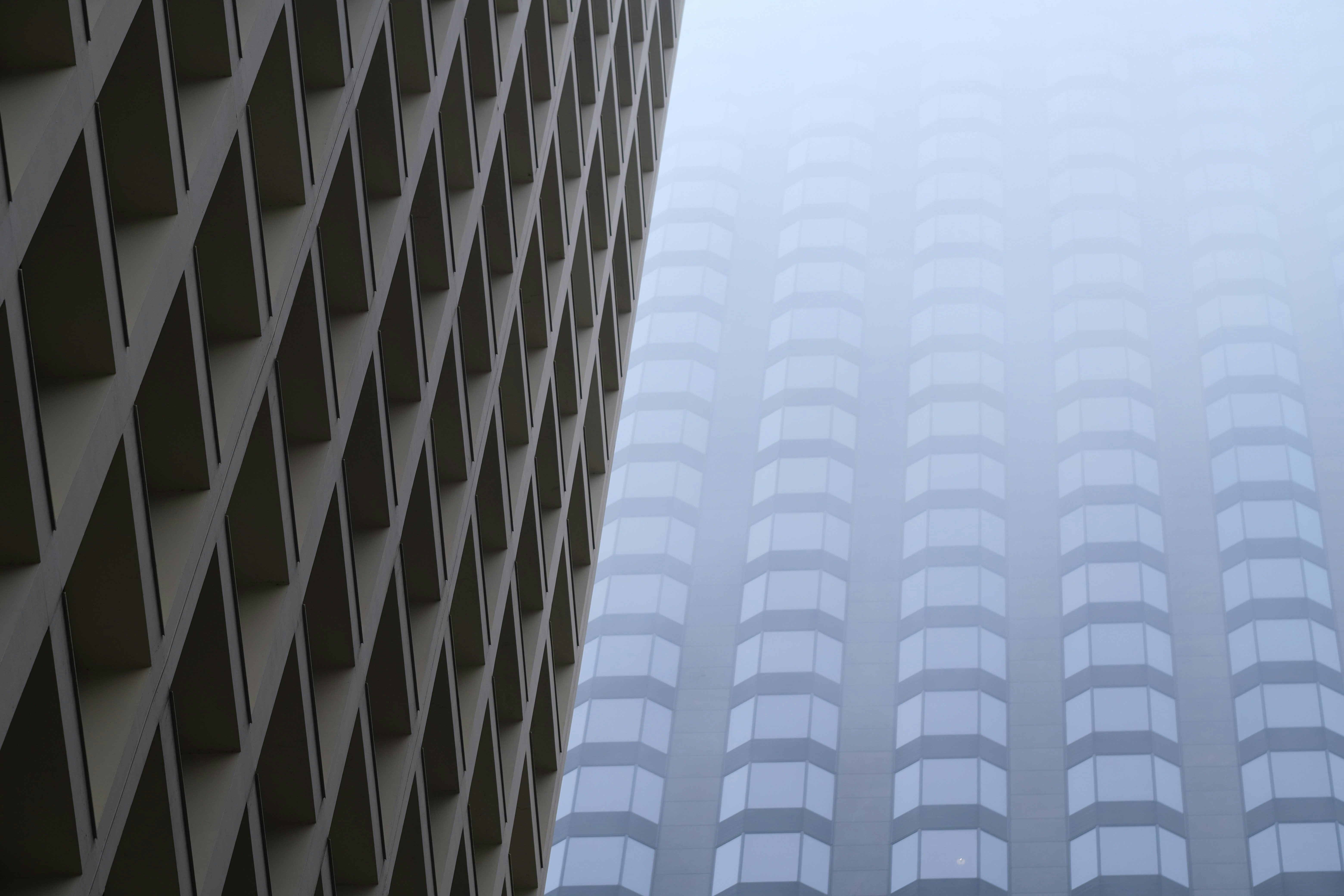 Two skyscrapers rise into fog, one with a pattern of square windows and the other with reflective glass panels.