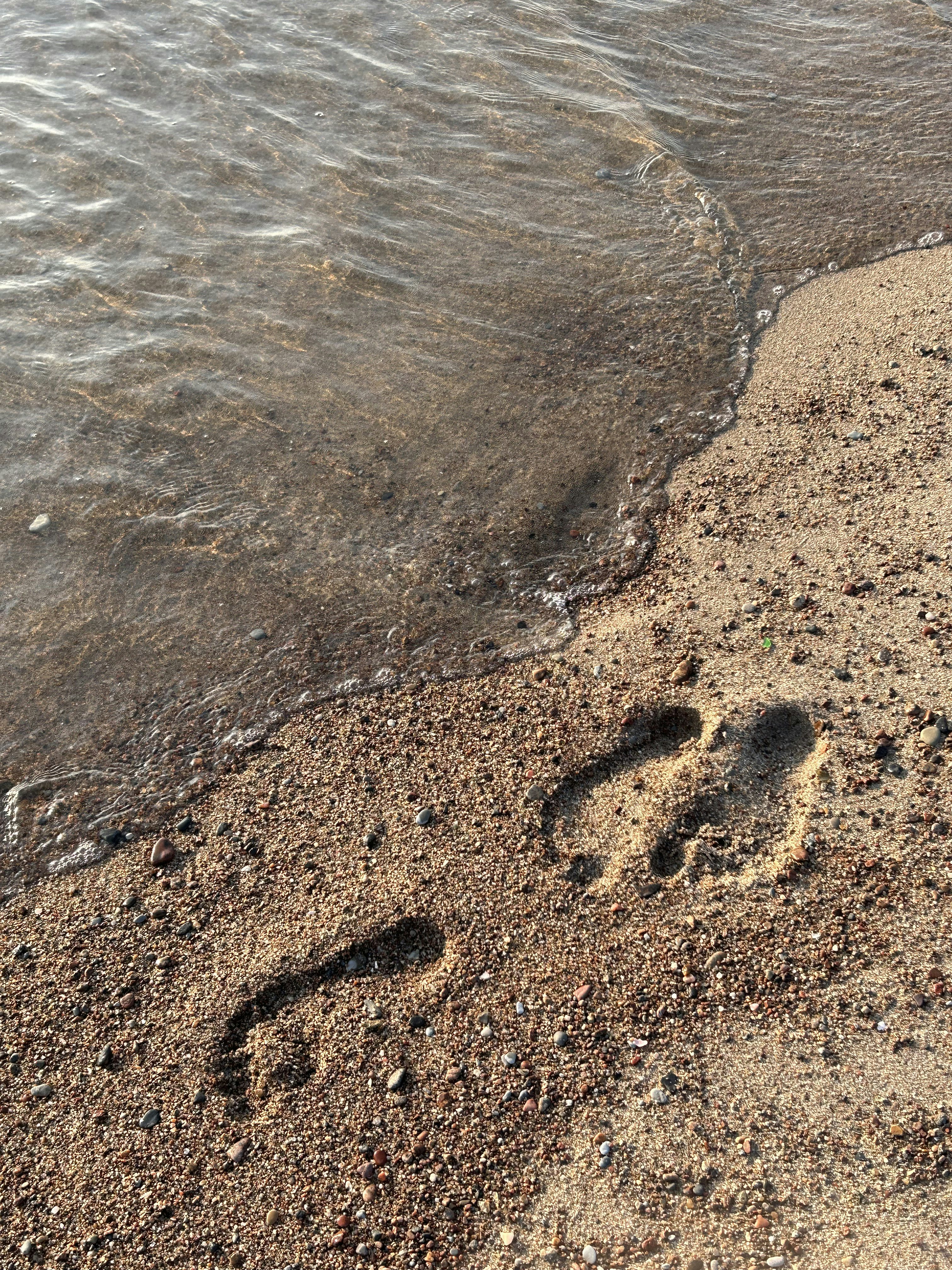 Footprints trail along damp sand at the water's edge as gentle waves wash over the shore.