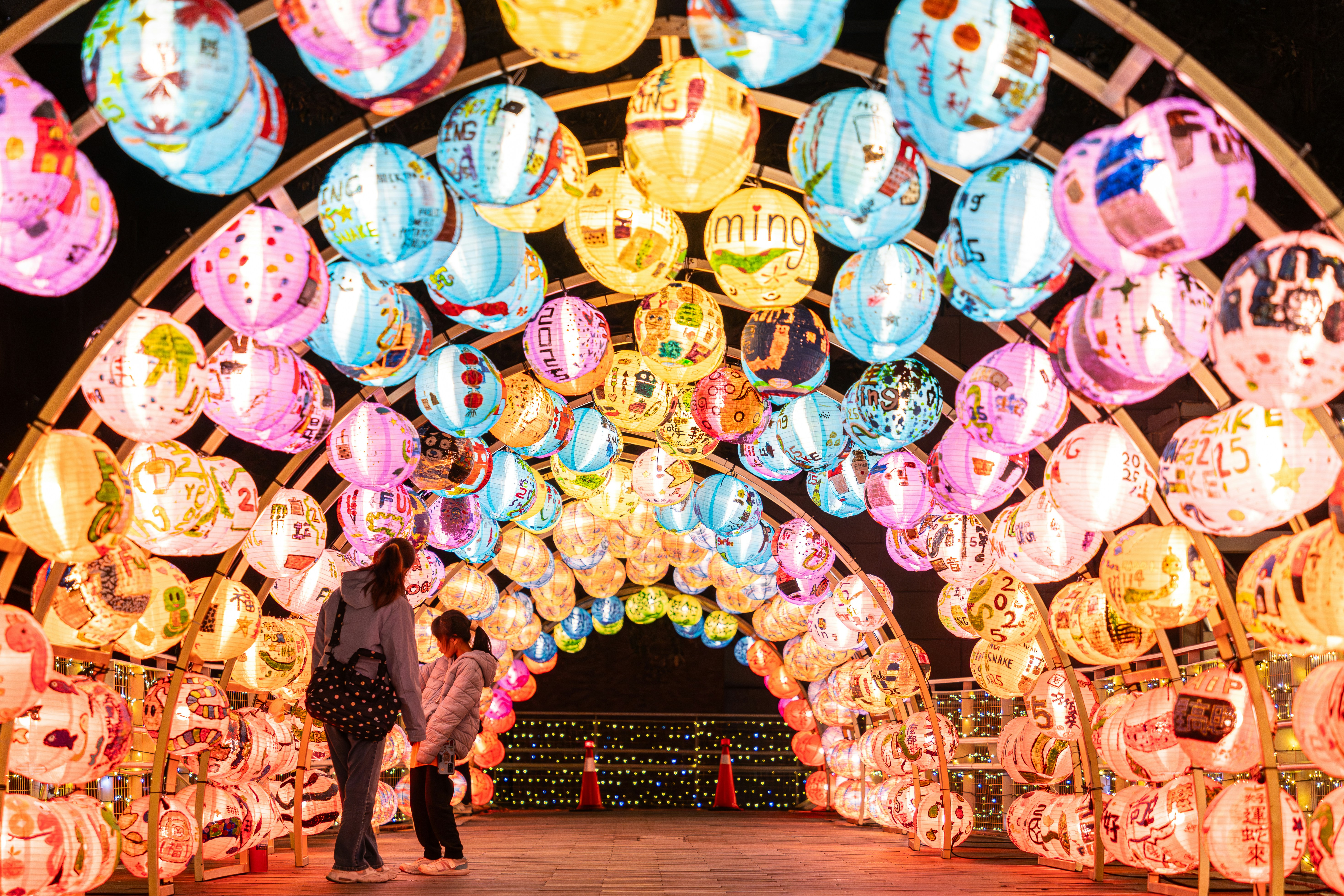 Colorful lantern tunnel glowing with blues, pinks, and yellows over two people walking beneath.