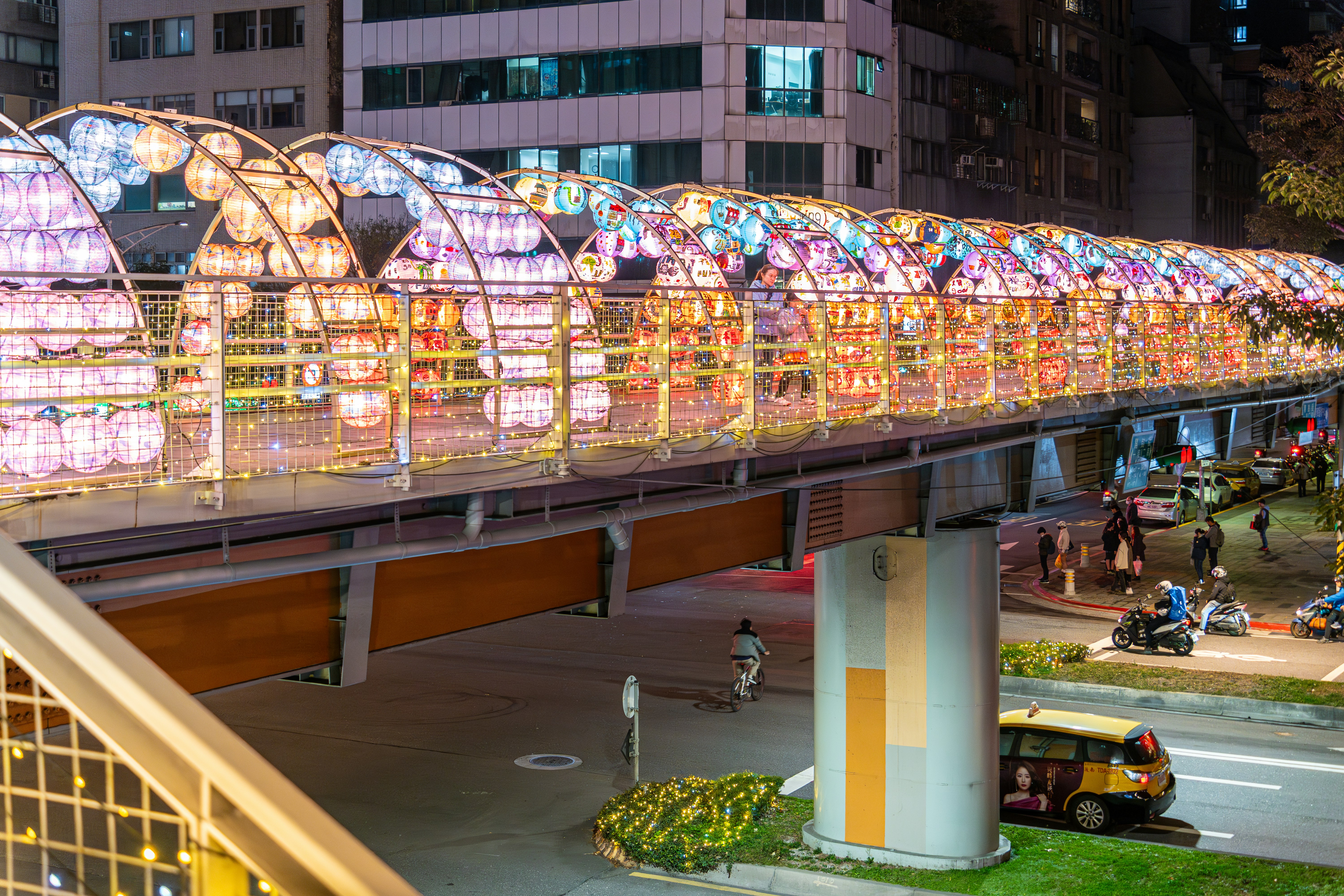 Colorful lanterns light up a pedestrian bridge in a cityscape, casting vibrant hues over the bustling street below.