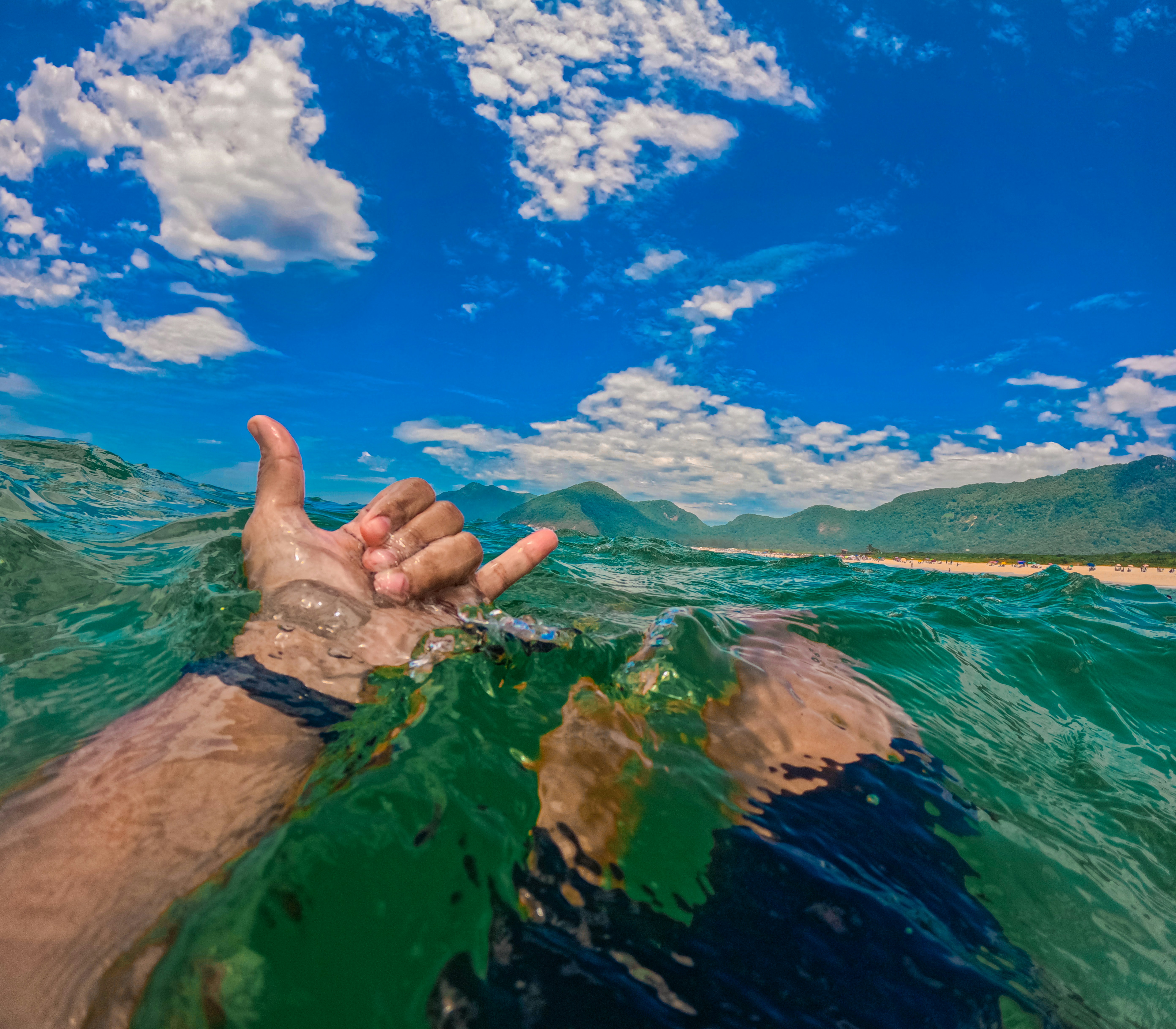 Hand making a shaka sign above vibrant green ocean waves under a bright blue sky with clouds.