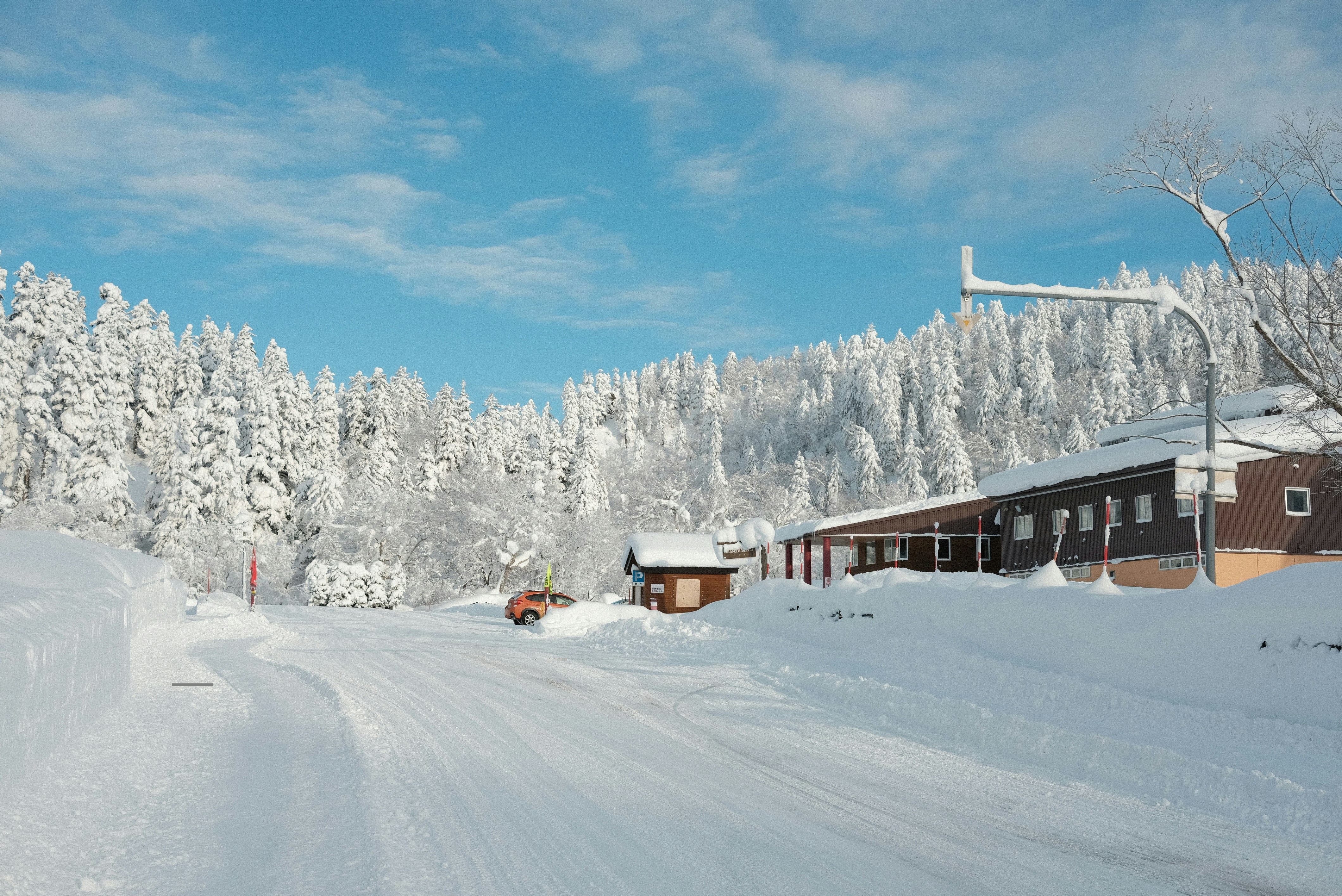 Snow-covered forest and rustic buildings with a bright orange vehicle under a clear blue sky.