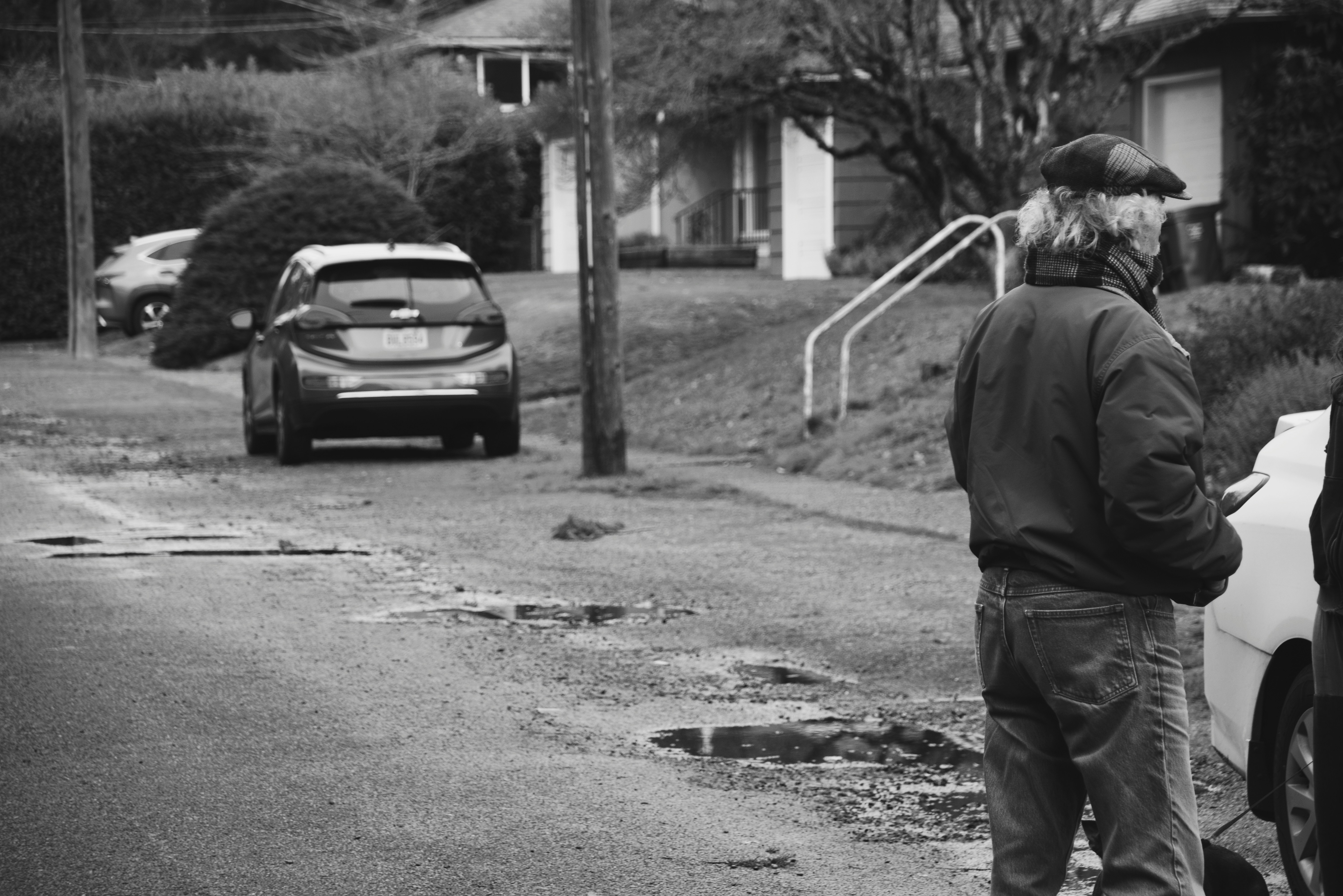 Man stands by a wet road with a parked car, framed by residential homes and overcast skies.