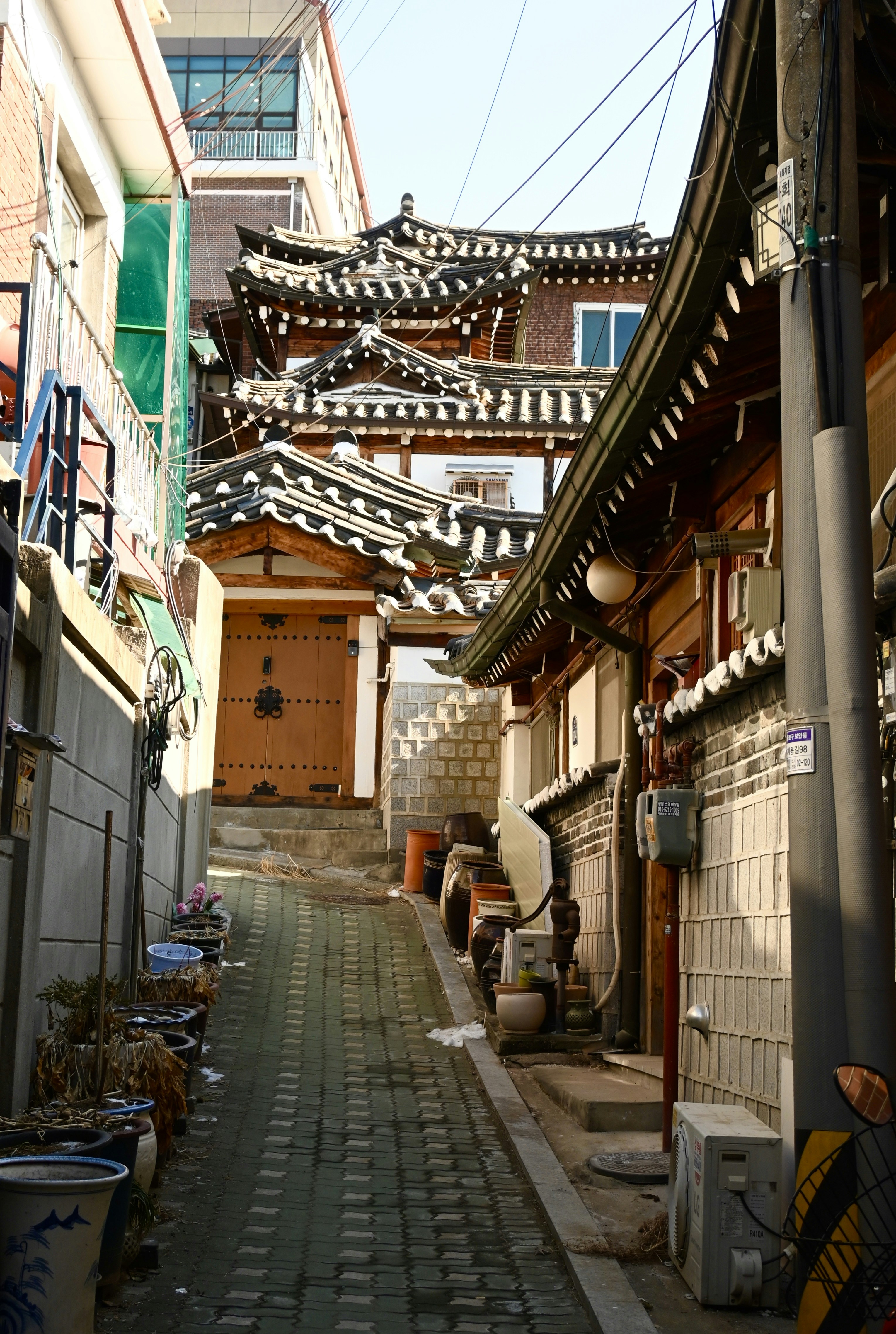 Narrow alleyway lined with traditional Korean houses, showcasing intricate roof designs and rustic charm. The path leads to a wooden gate, inviting exploration.