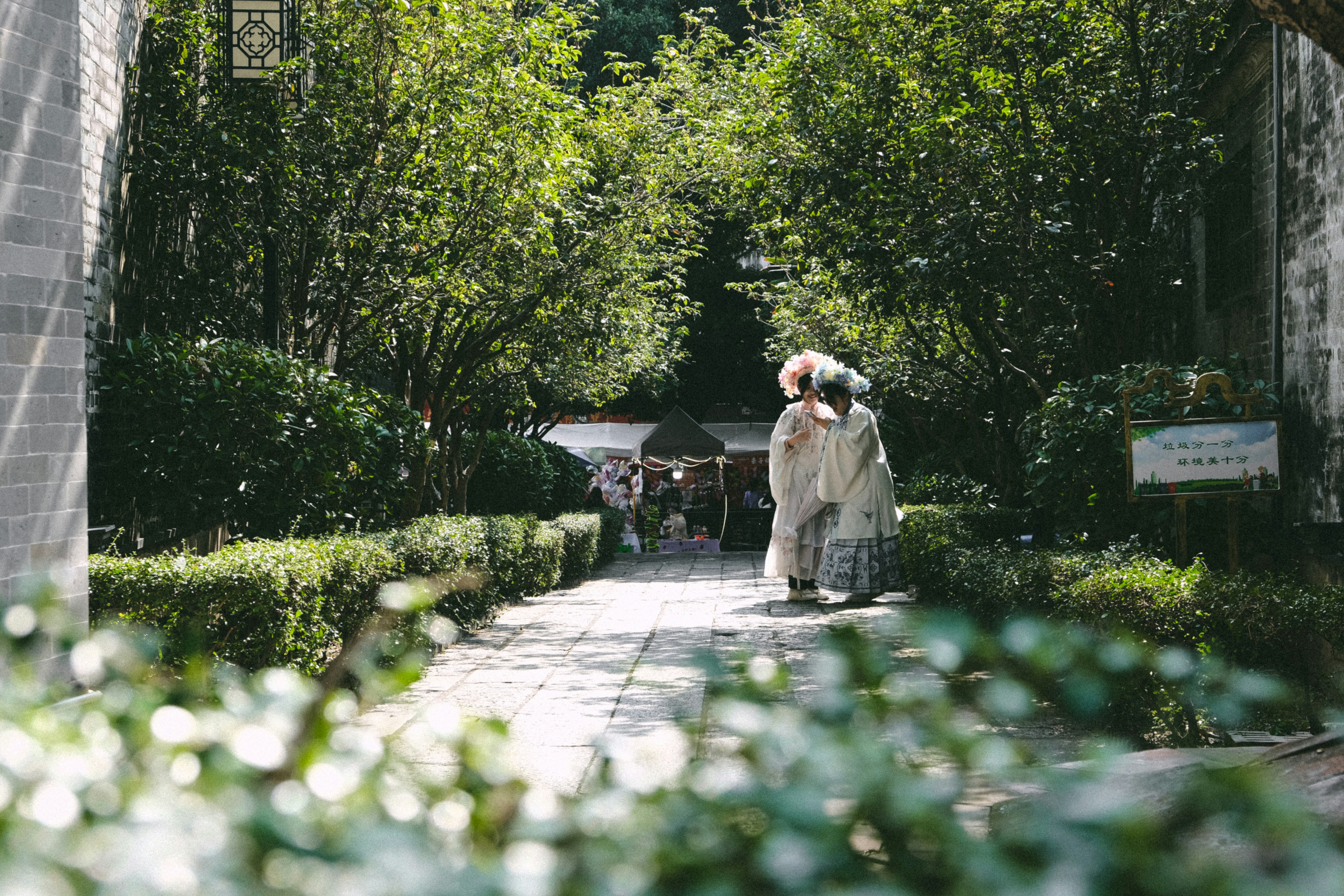 Two figures in traditional attire stroll beneath arching trees, surrounded by lush greenery on a sunlit garden path.