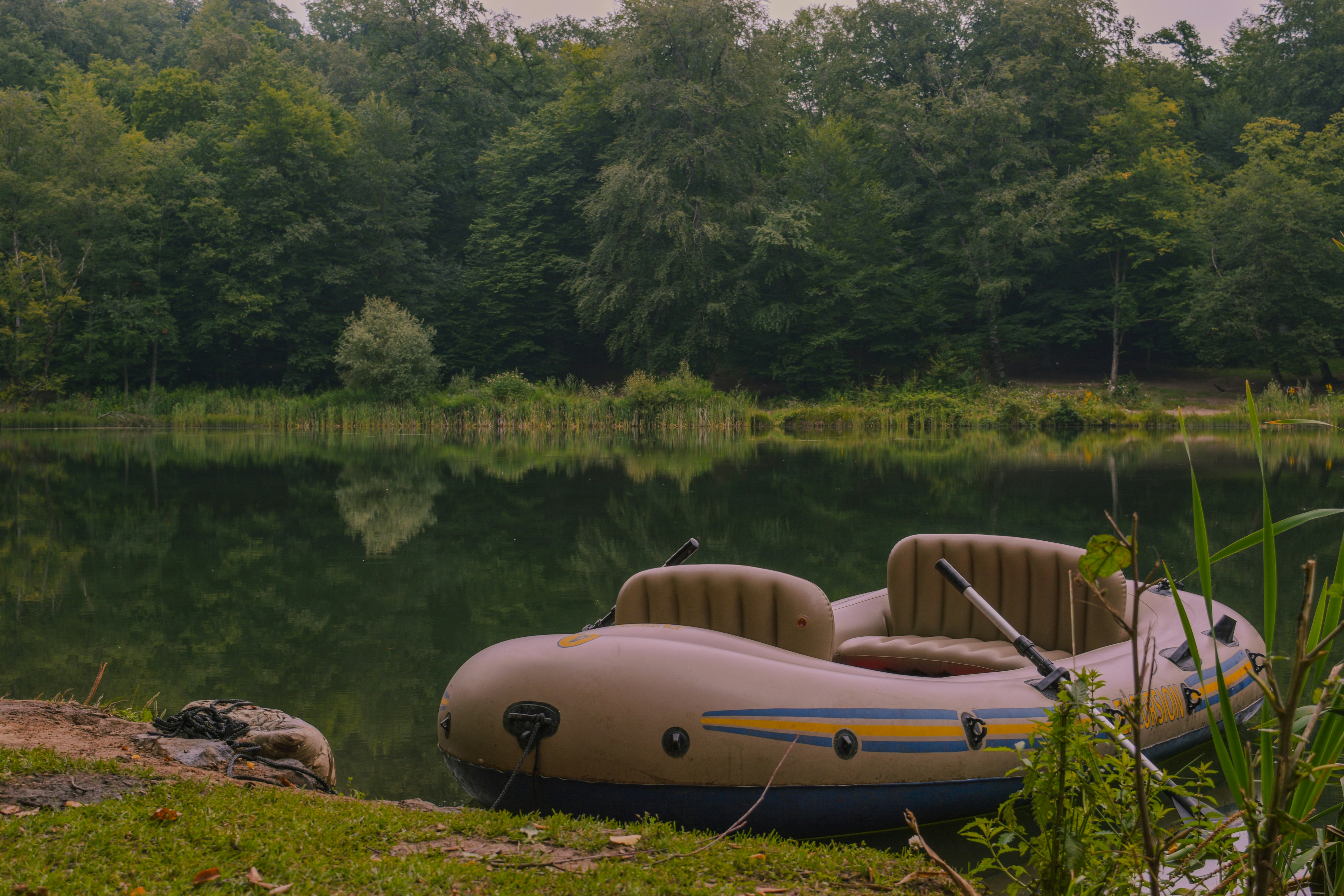 Inflatable boat rests quietly by a tranquil lake, surrounded by dense forest greenery with calm waters reflecting the scene.
