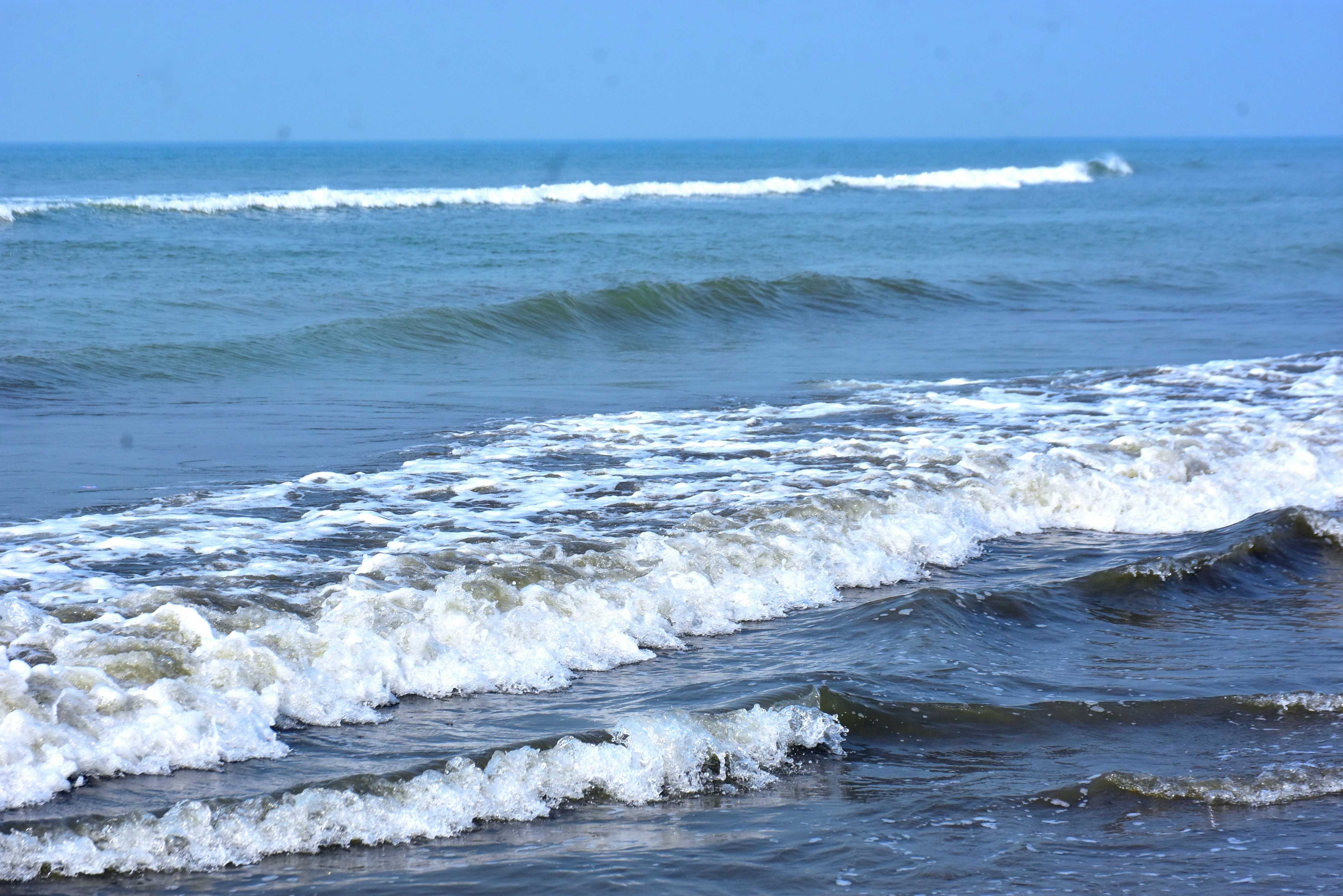 Gentle waves roll onto the shore under a clear blue sky, with frothy crests contrasting against deep blue and green hues.
