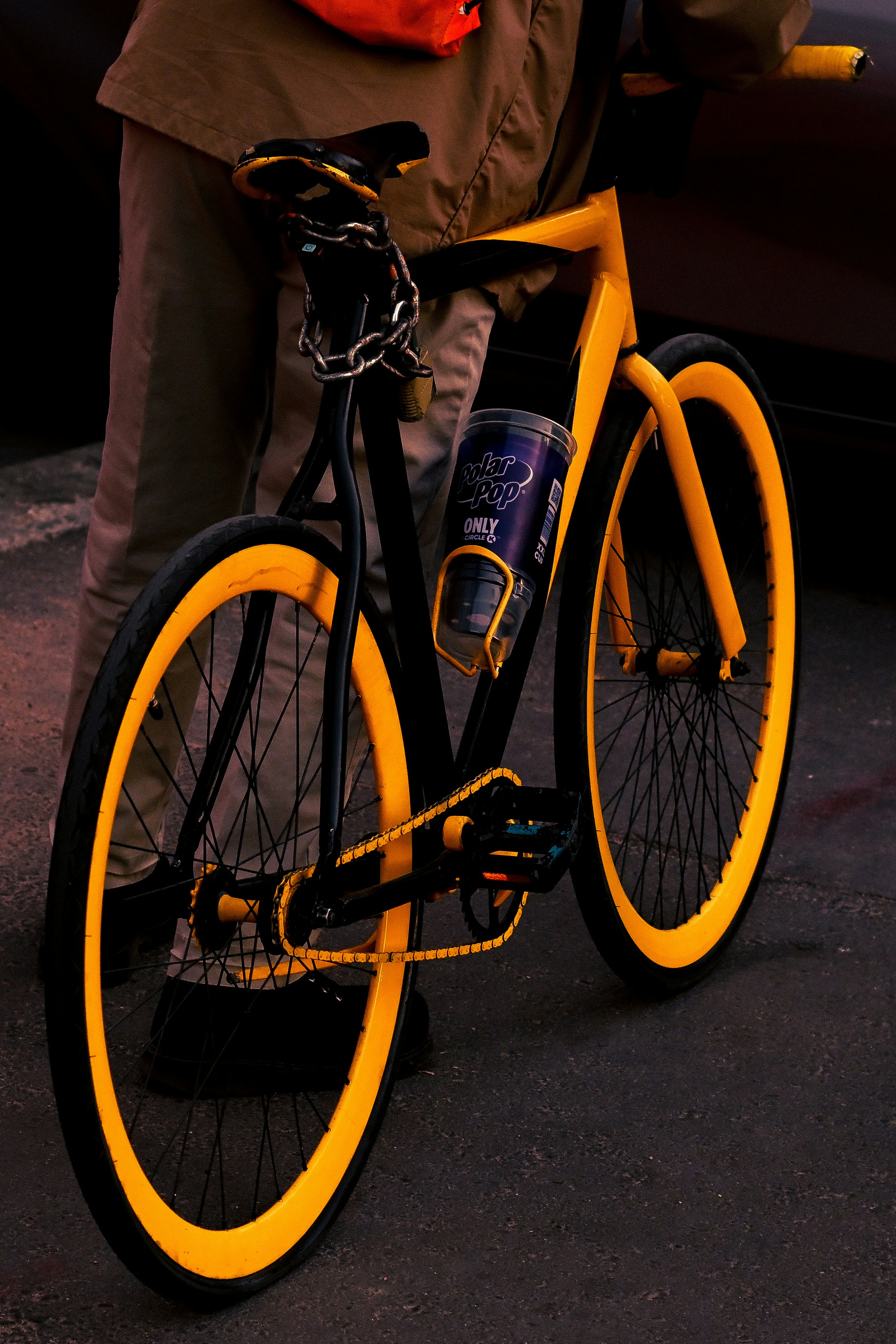 A yellow bicycle with bright orange rims and a mounted water bottle sits on a dark street, with a rider nearby.