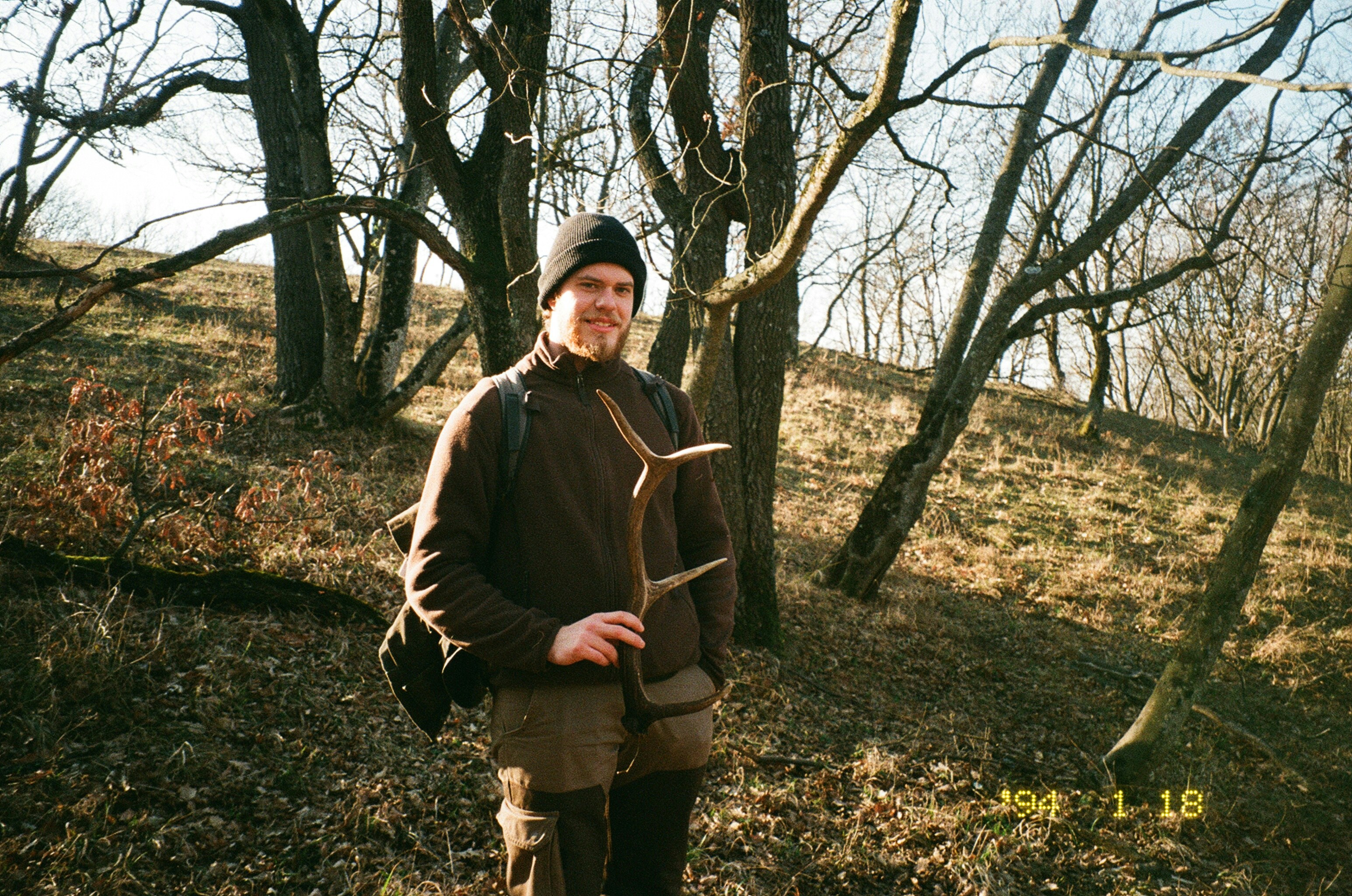 Man in earthy clothing holds an antler amidst a sunlit forest, with bare trees and golden afternoon light creating a serene atmosphere.
