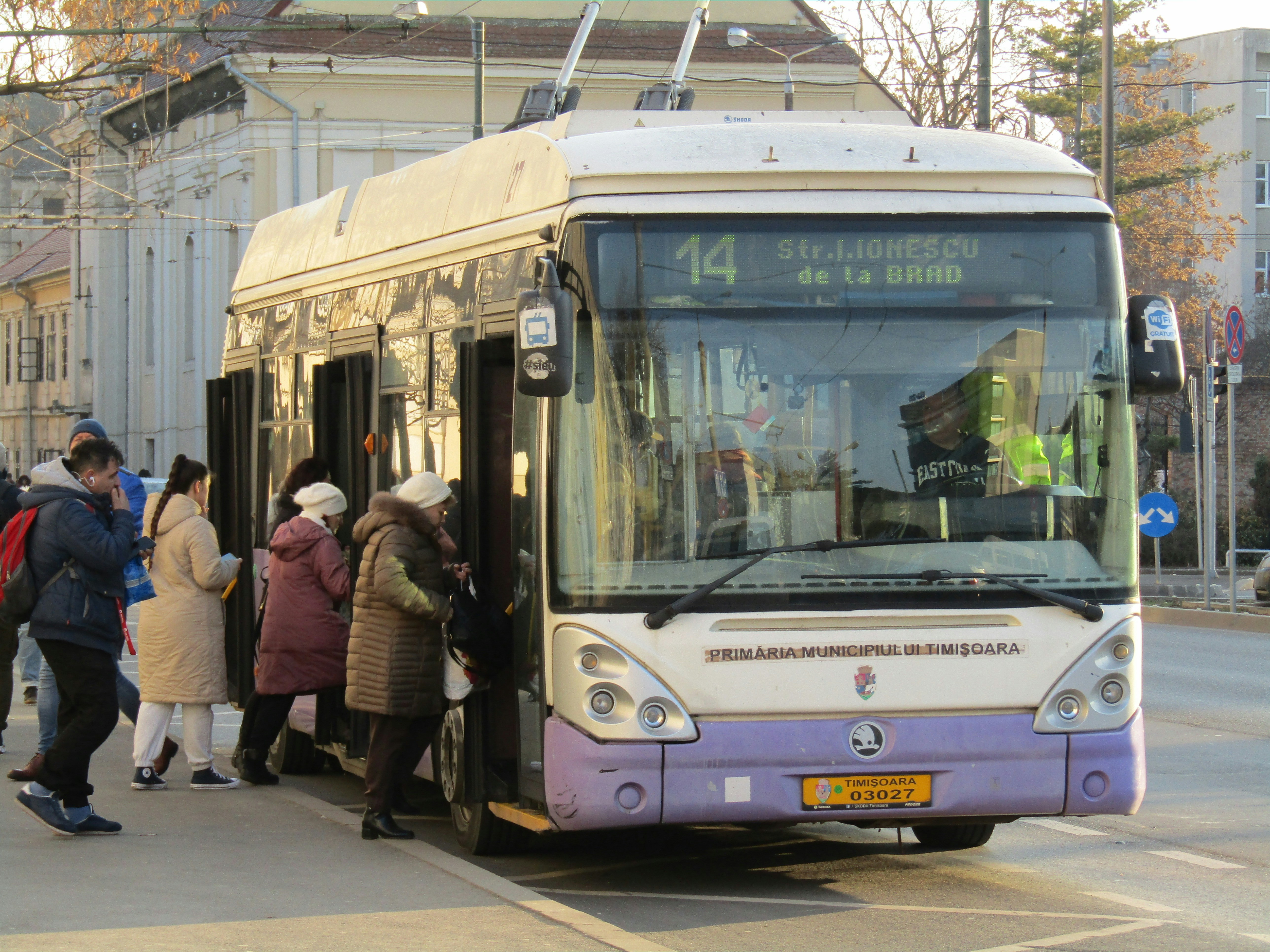 People board a lavender-and-white city bus at a busy street stop, route 14 clearly visible on the front.