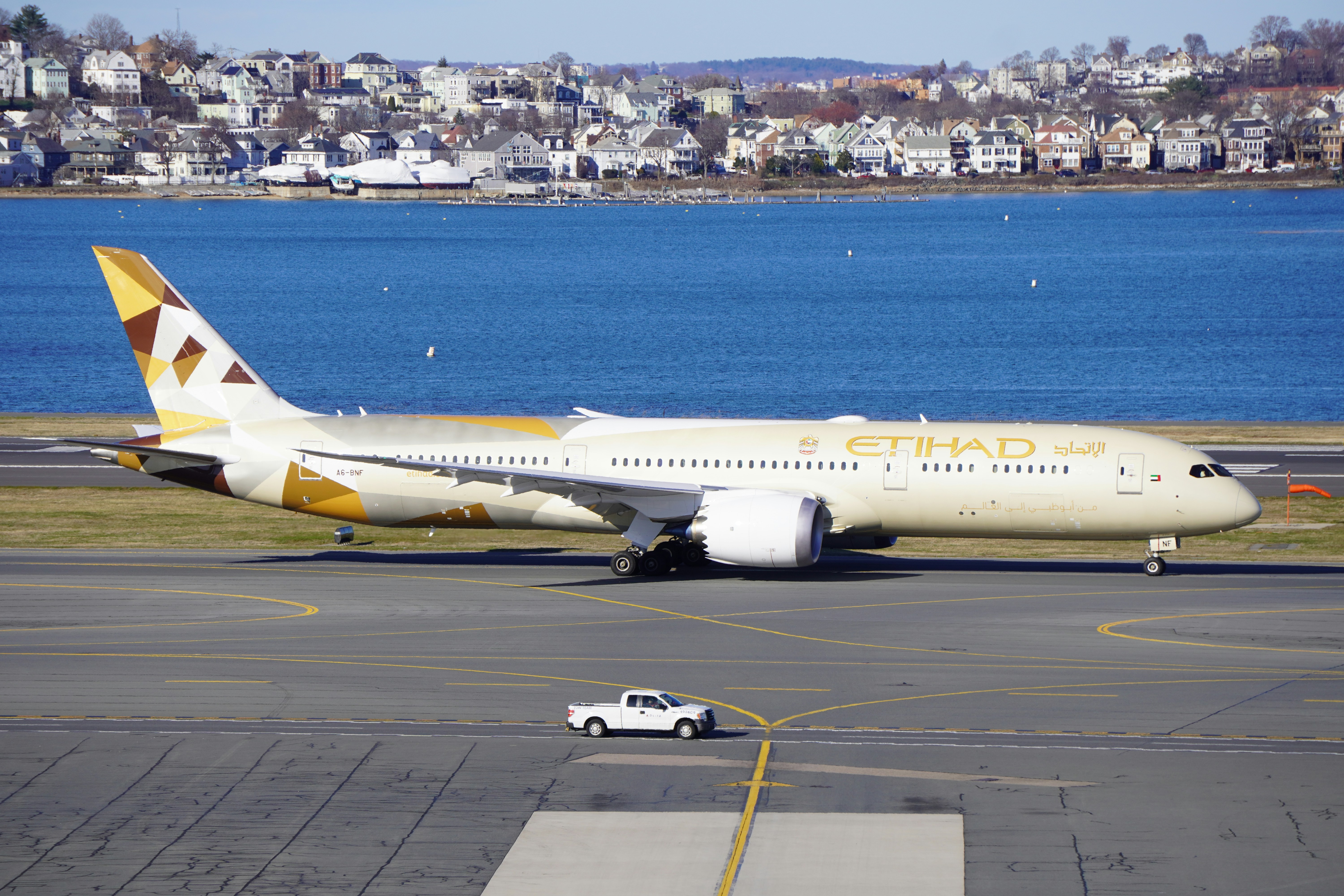 Passenger airplane with golden accents taxiing on a runway beside a blue waterfront.