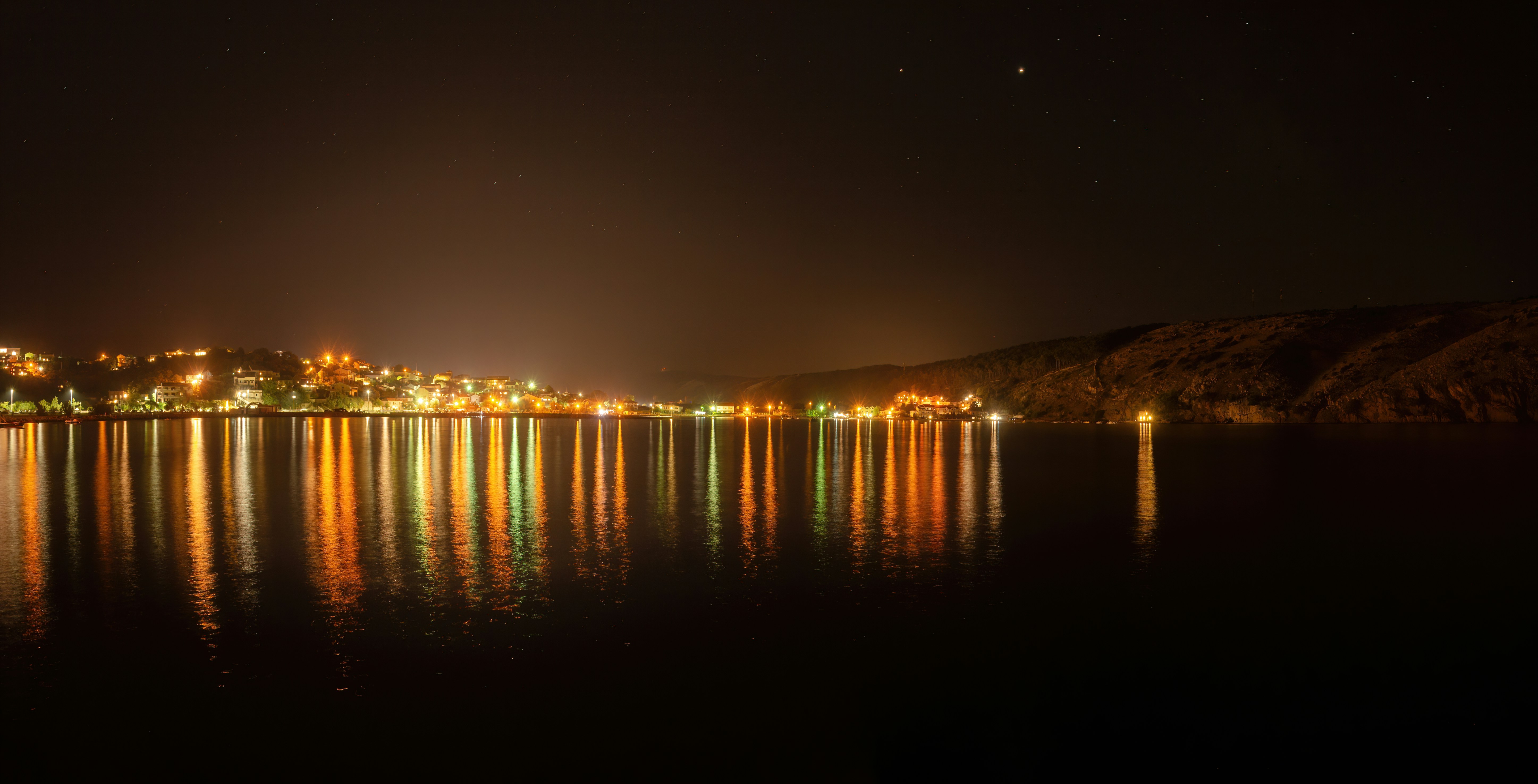 City lights shimmer across a tranquil bay under a starry night sky, casting colorful reflections on the water.