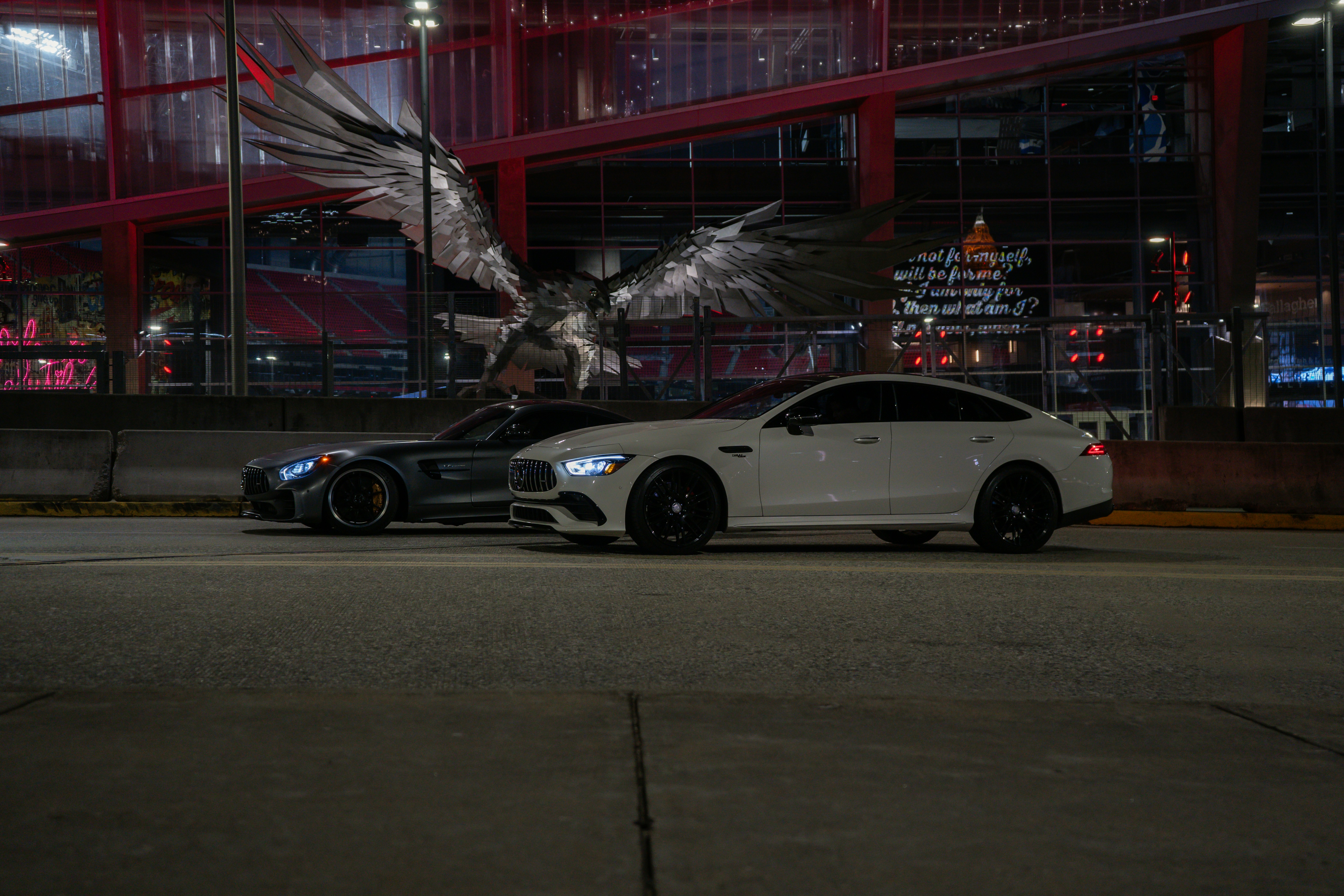 Two luxury cars parked under a winged sculpture in a city at night, highlighted by streetlights and reflections from nearby buildings.