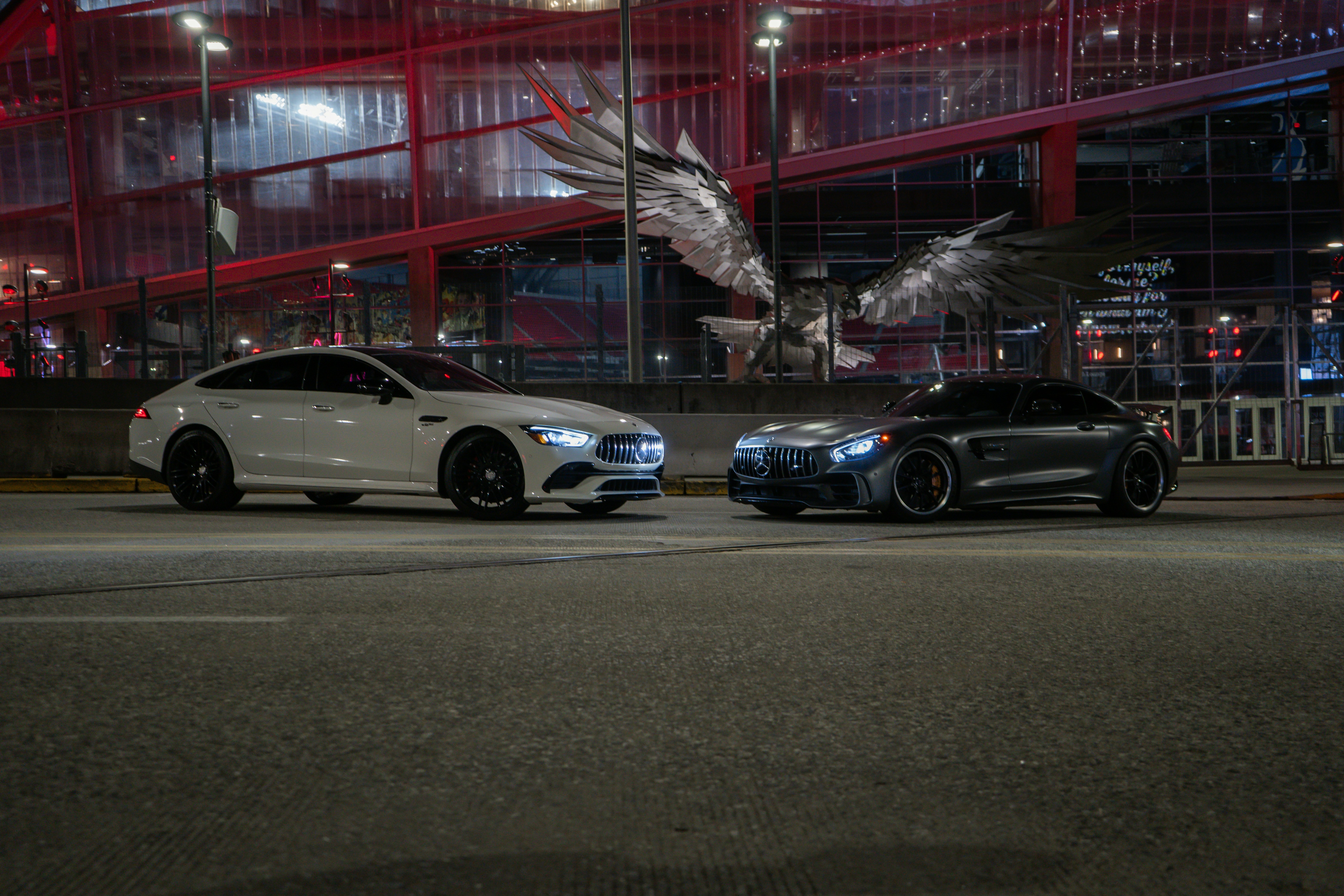 Luxury cars in white and metallic gray parked under a winged sculpture against a red-lit modern glass building.
