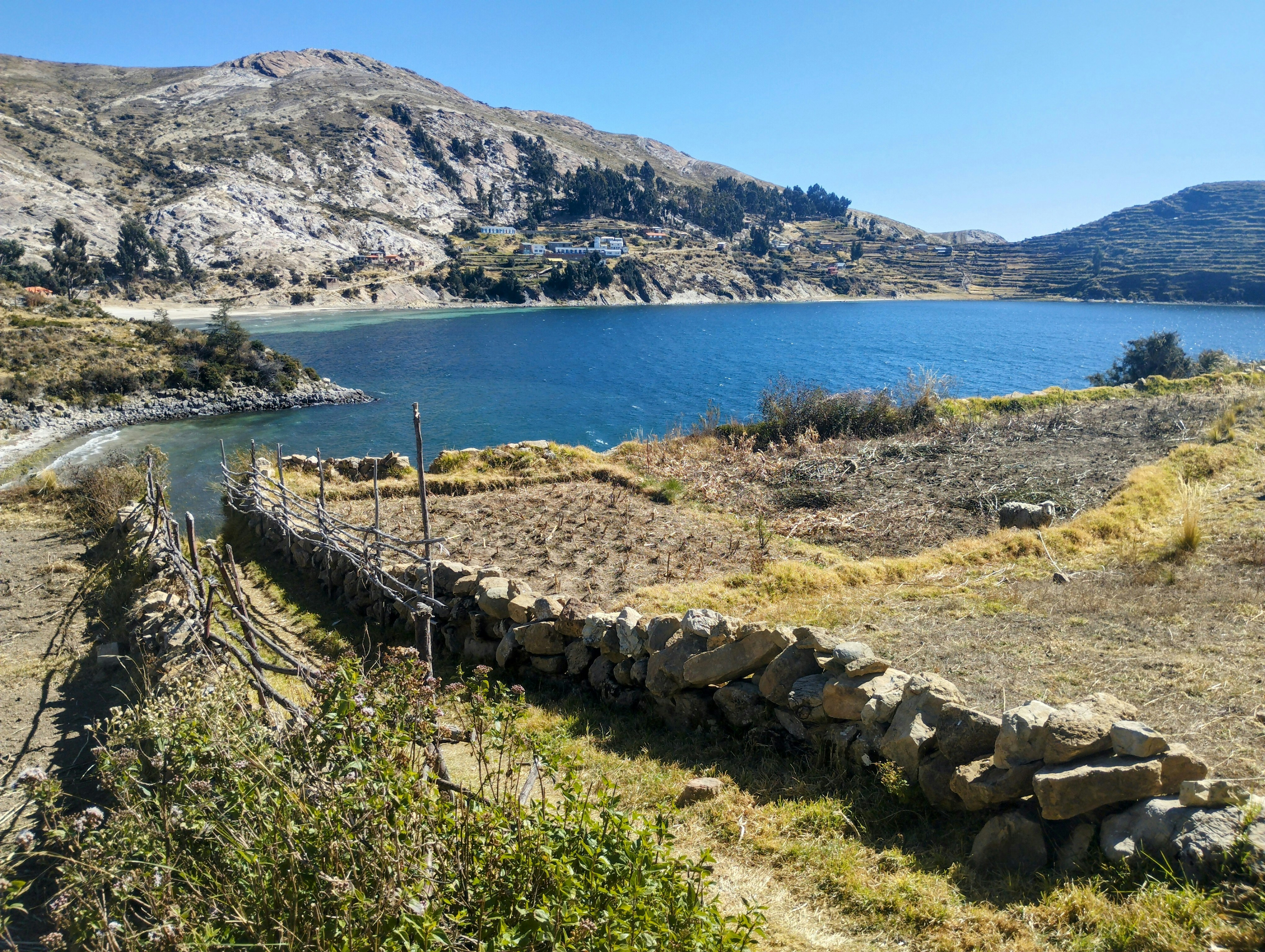 Rugged landscape with a stone-walled path leading toward a blue lake, framed by sunlit hills under a clear sky.
