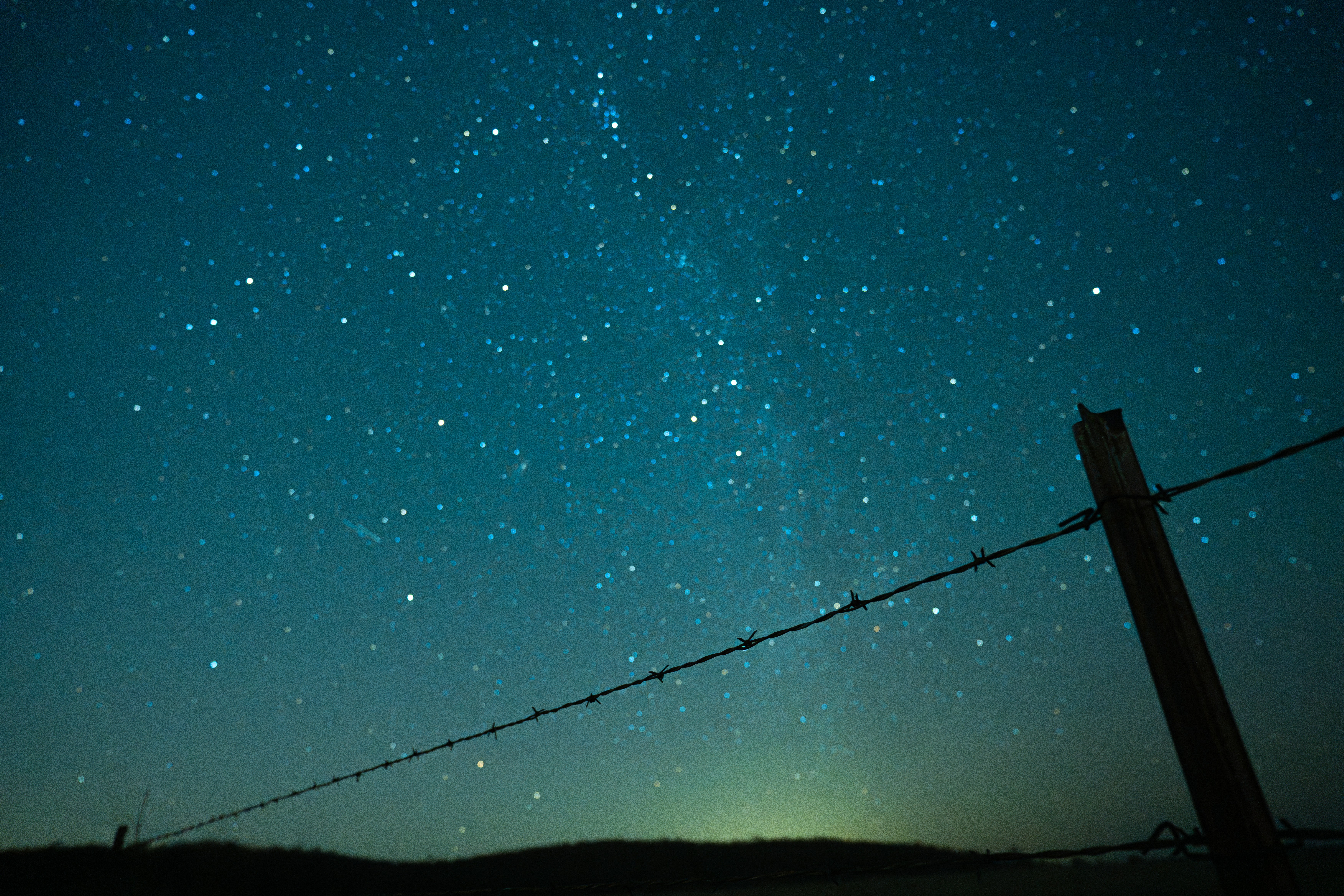 Star-filled night sky over a silhouetted barbed wire fence with a faint horizon glow.