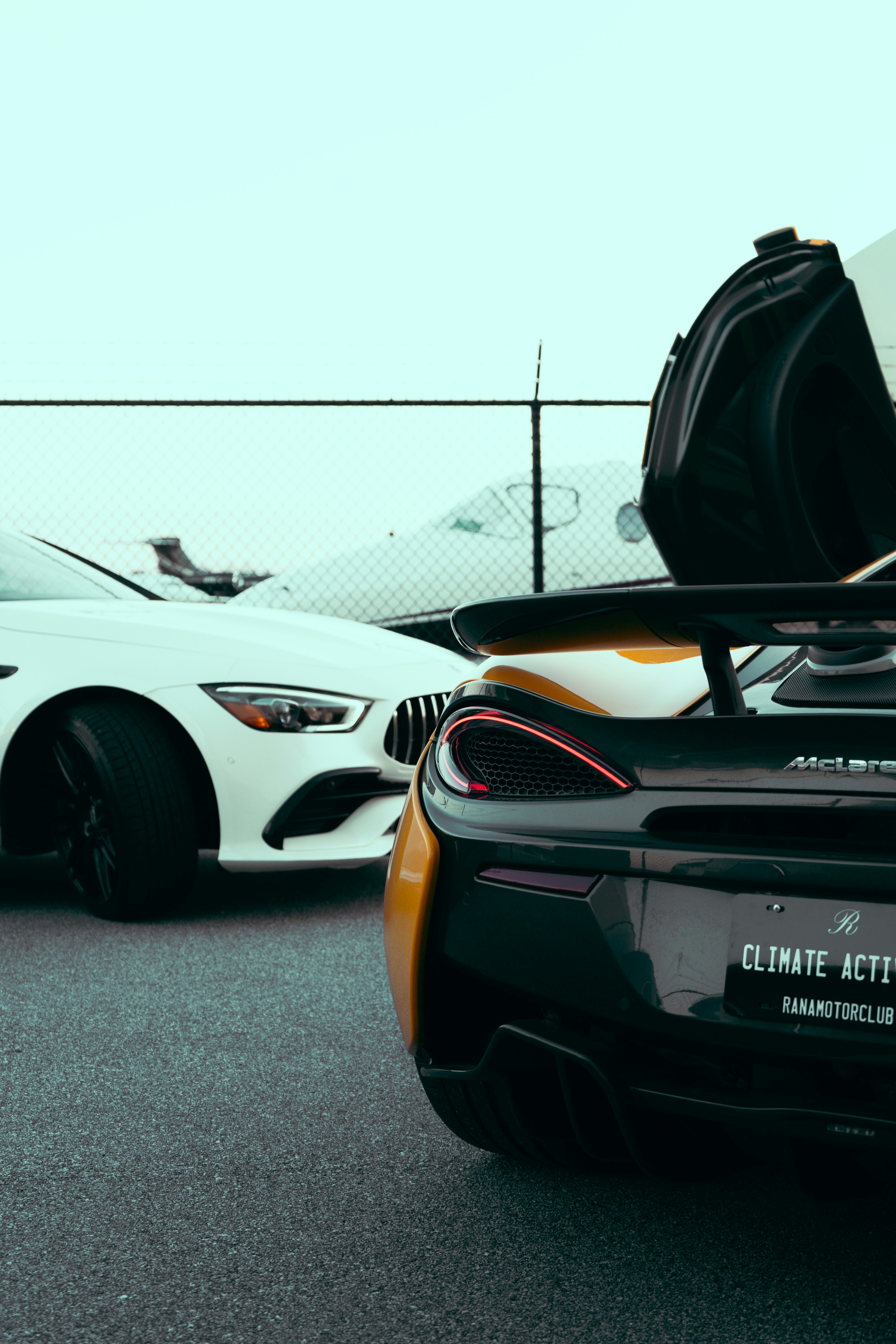 Rear-Quarter Spotlight: McLaren vs. White BMW Framed by a Chain-Link Horizon